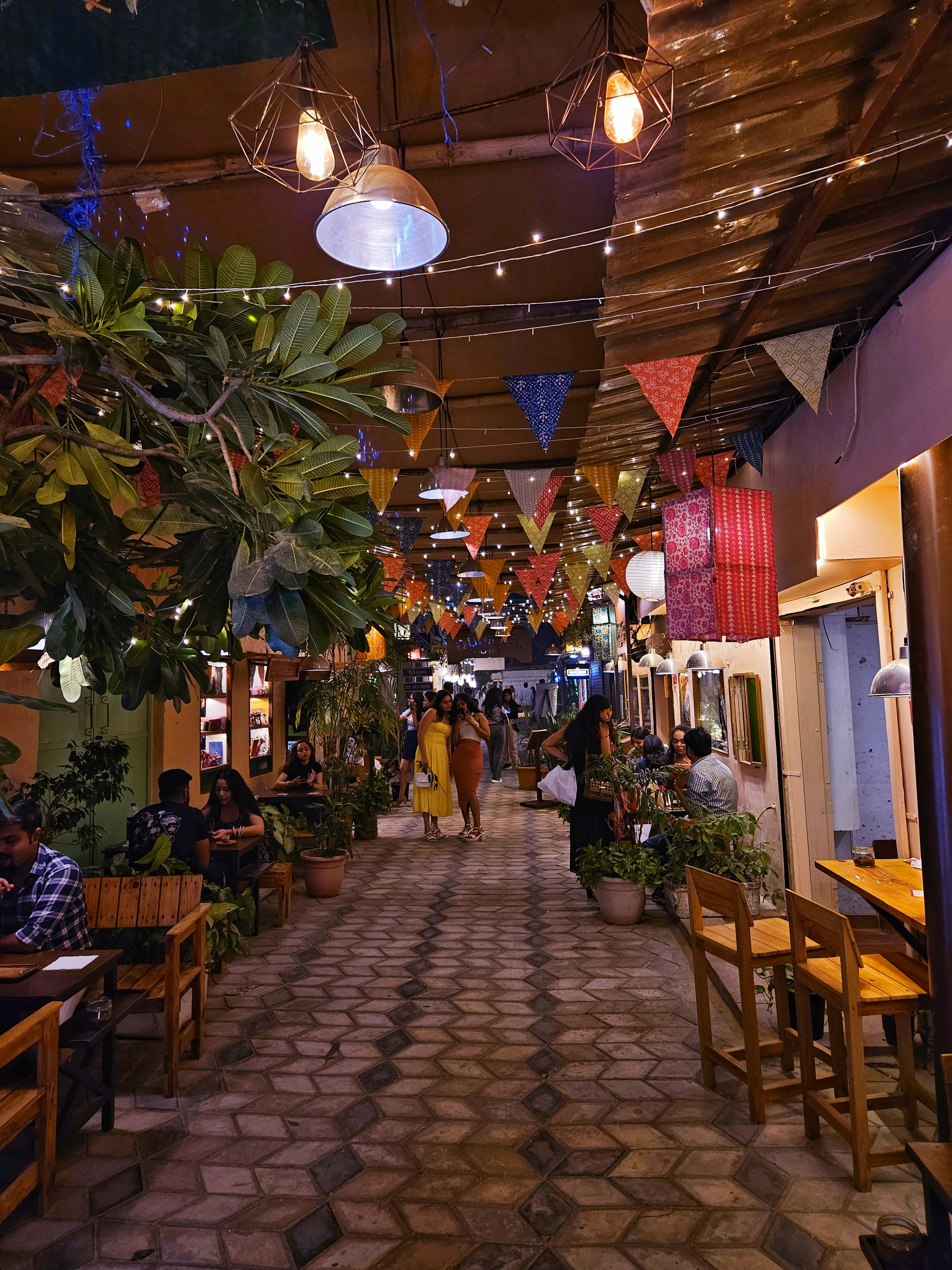 A night-time photograph of a plant-lined alleyway café decorated with bunting and string lights, with patrons seated at wooden tables.