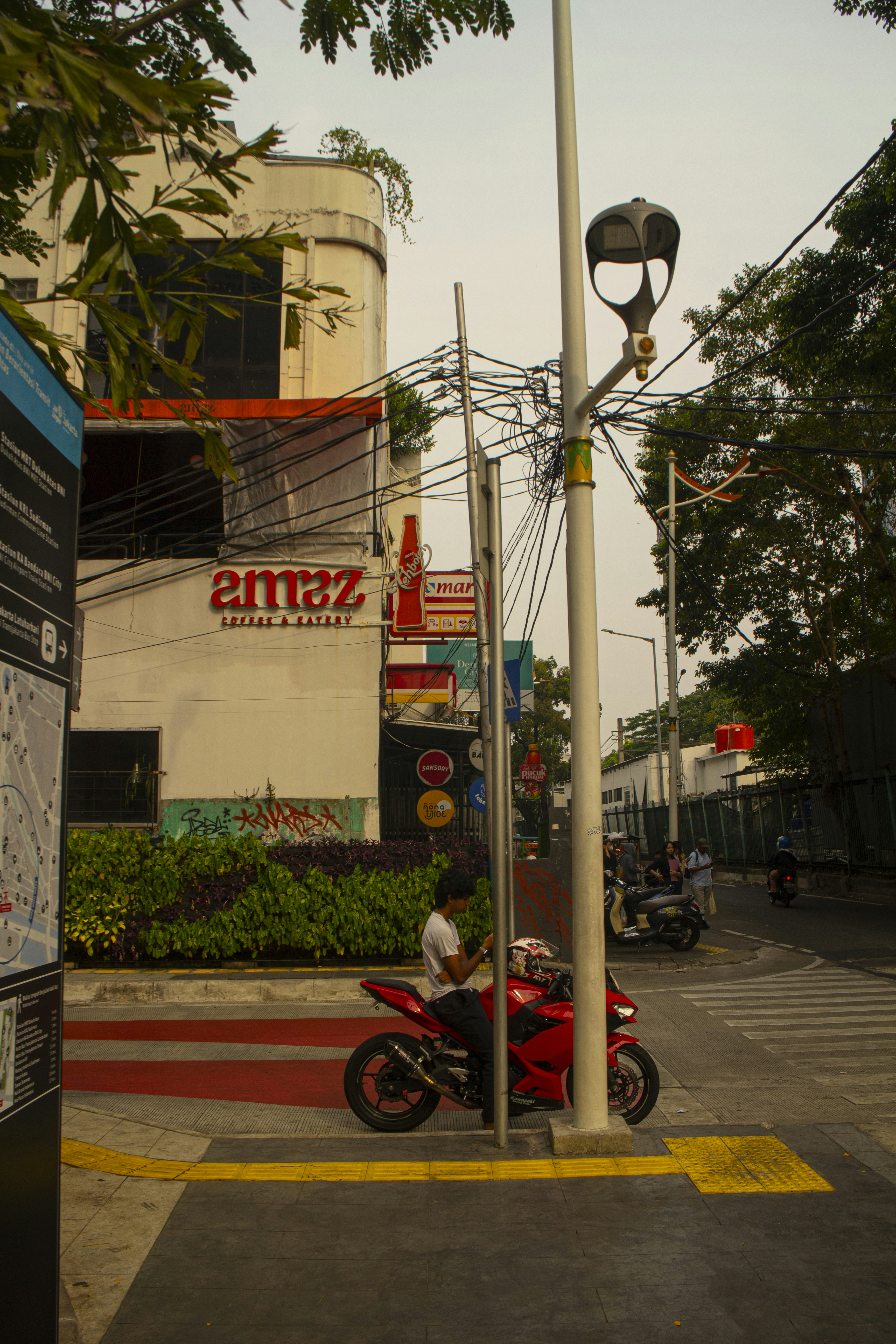 Crimson sportbike parked at a crosswalk beside a streetlamp, with a man seated nearby and storefronts in the background. Overhead wires crisscross, underscoring the bustling city scene.