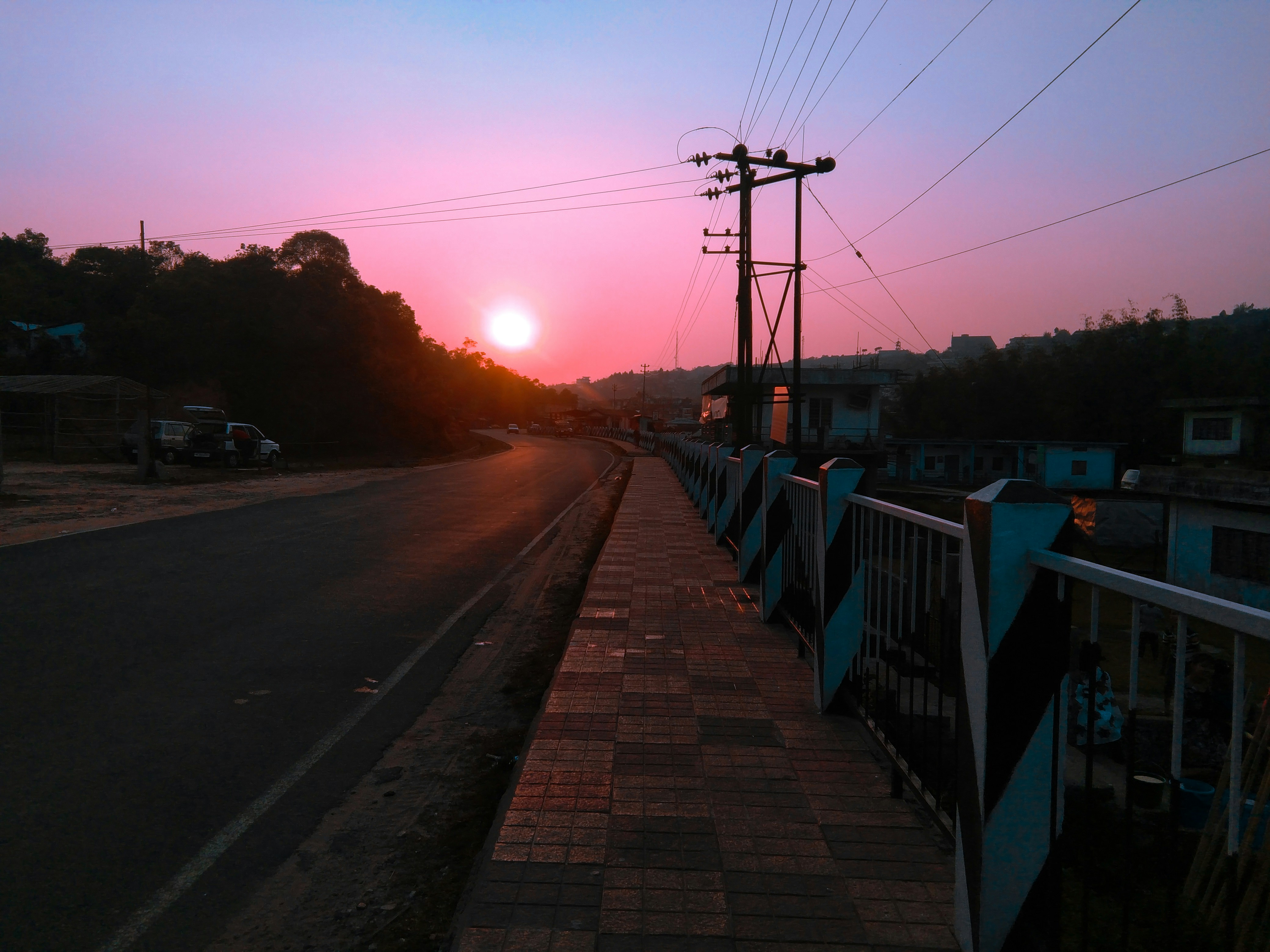 Sunset casts a warm glow over a quiet city street lined with trees and power lines.