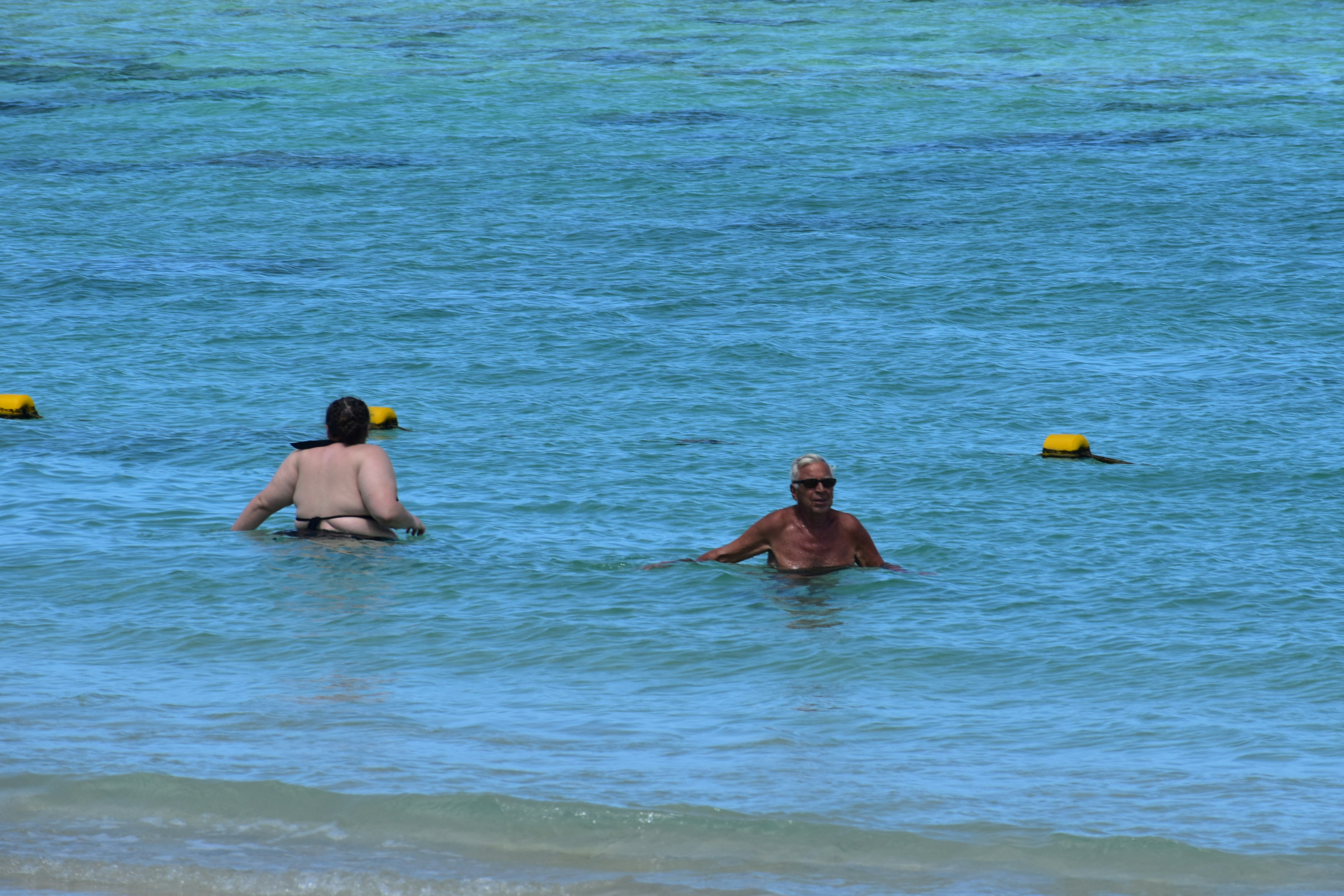 Two people enjoying a leisurely swim in clear blue sea with floating buoys nearby.