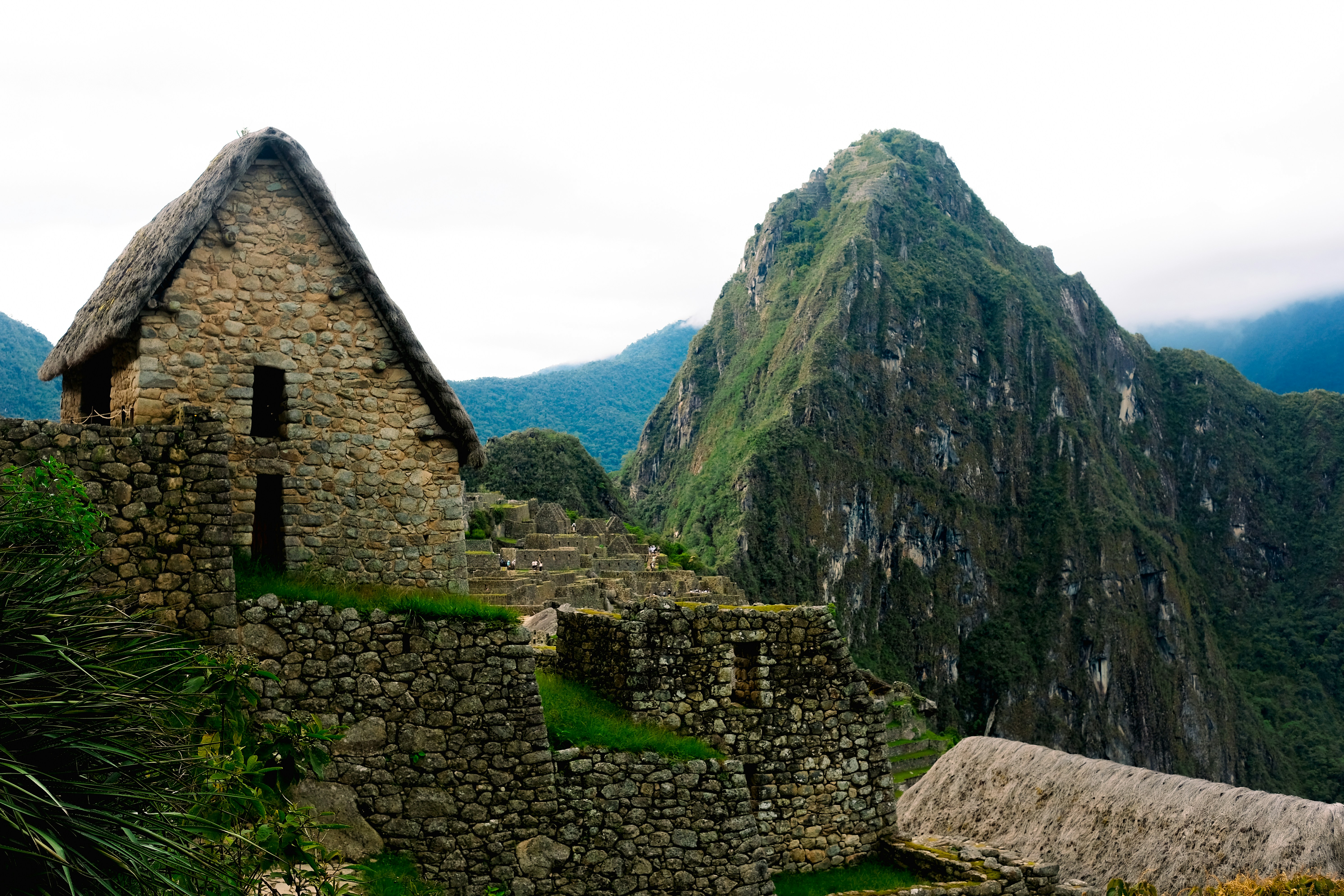 Rustic stone house with a thatched roof framed by a lush green mountain backdrop under an overcast sky.