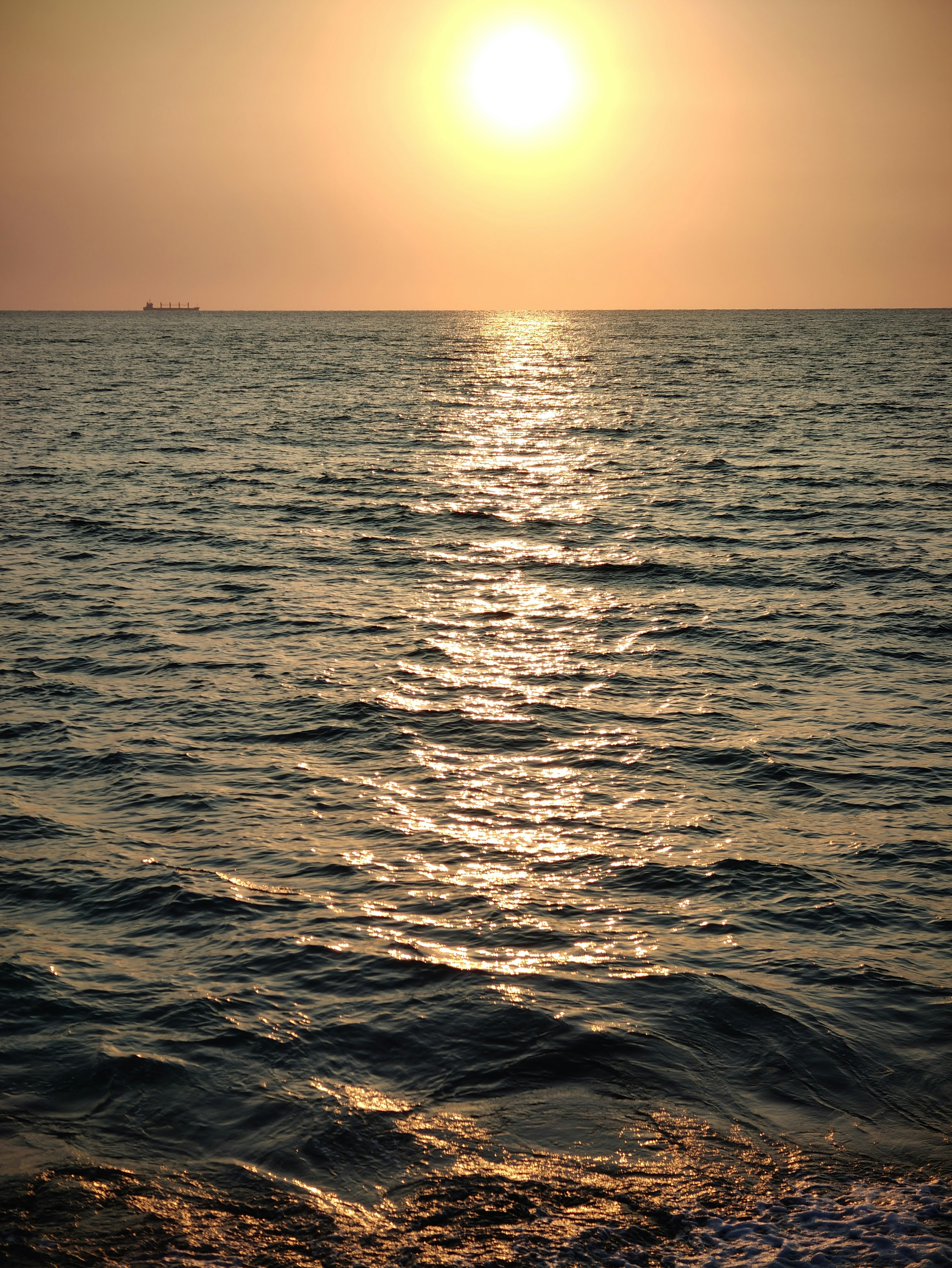Sunset over the ocean, with shimmering reflections on the water's surface and a distant silhouette of a ship. 
