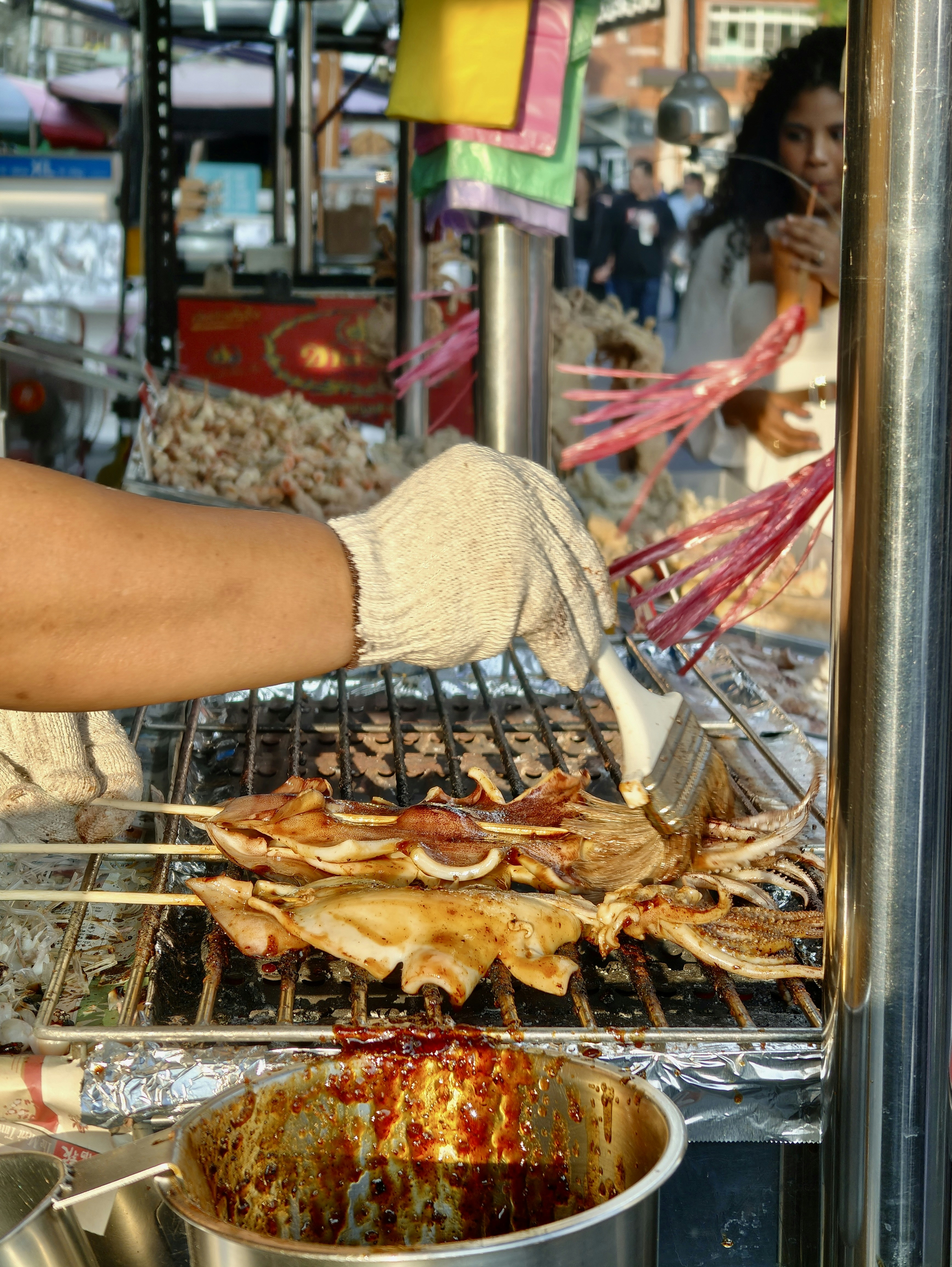 Grilled skewers of seafood being brushed with sauce at a bustling street food stall, with a customer in the background enjoying a drink.