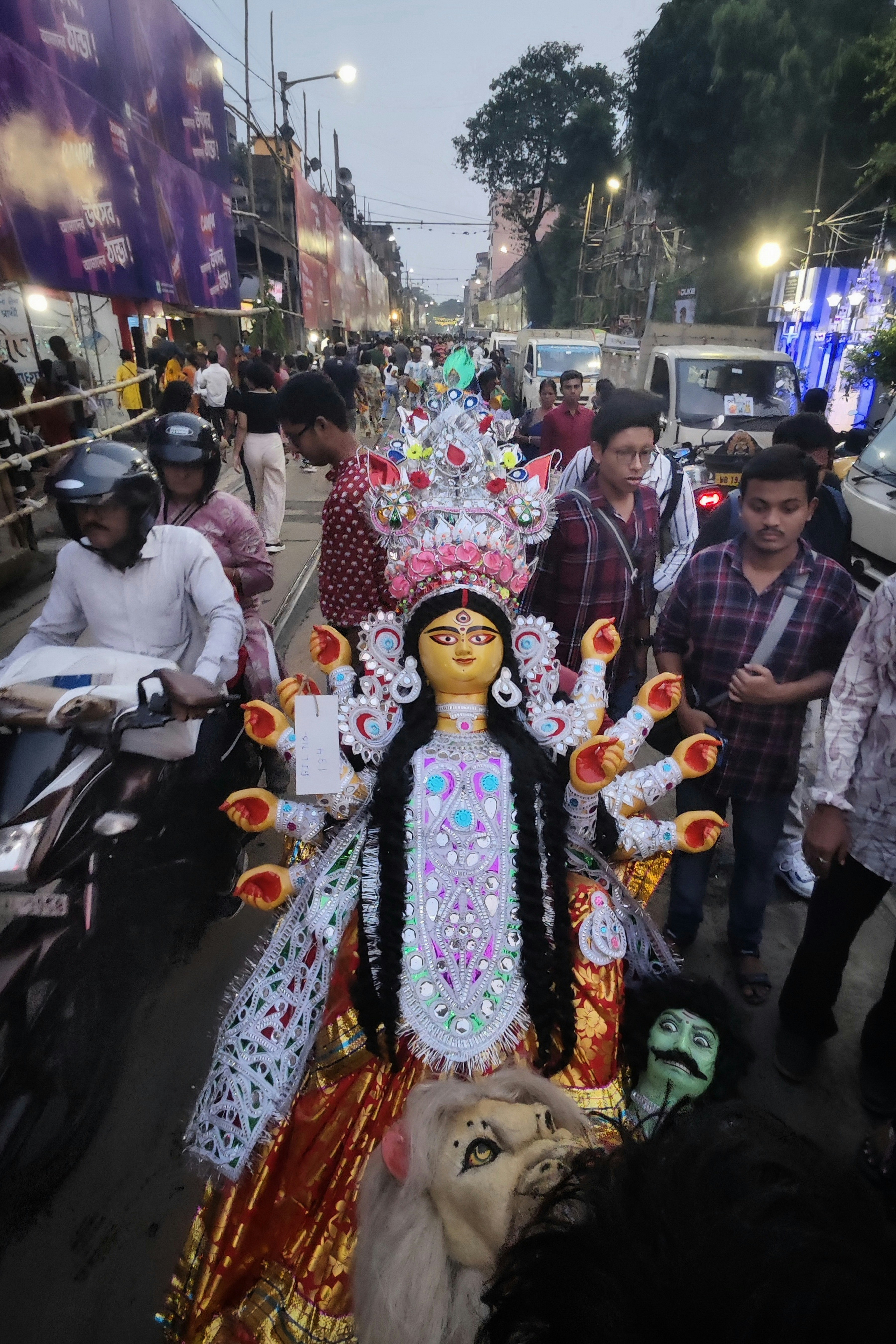 A photograph of a colorfully adorned Durga idol leading a crowded street procession, surrounded by spectators, scooters, and pedestrians as dusk light settles.