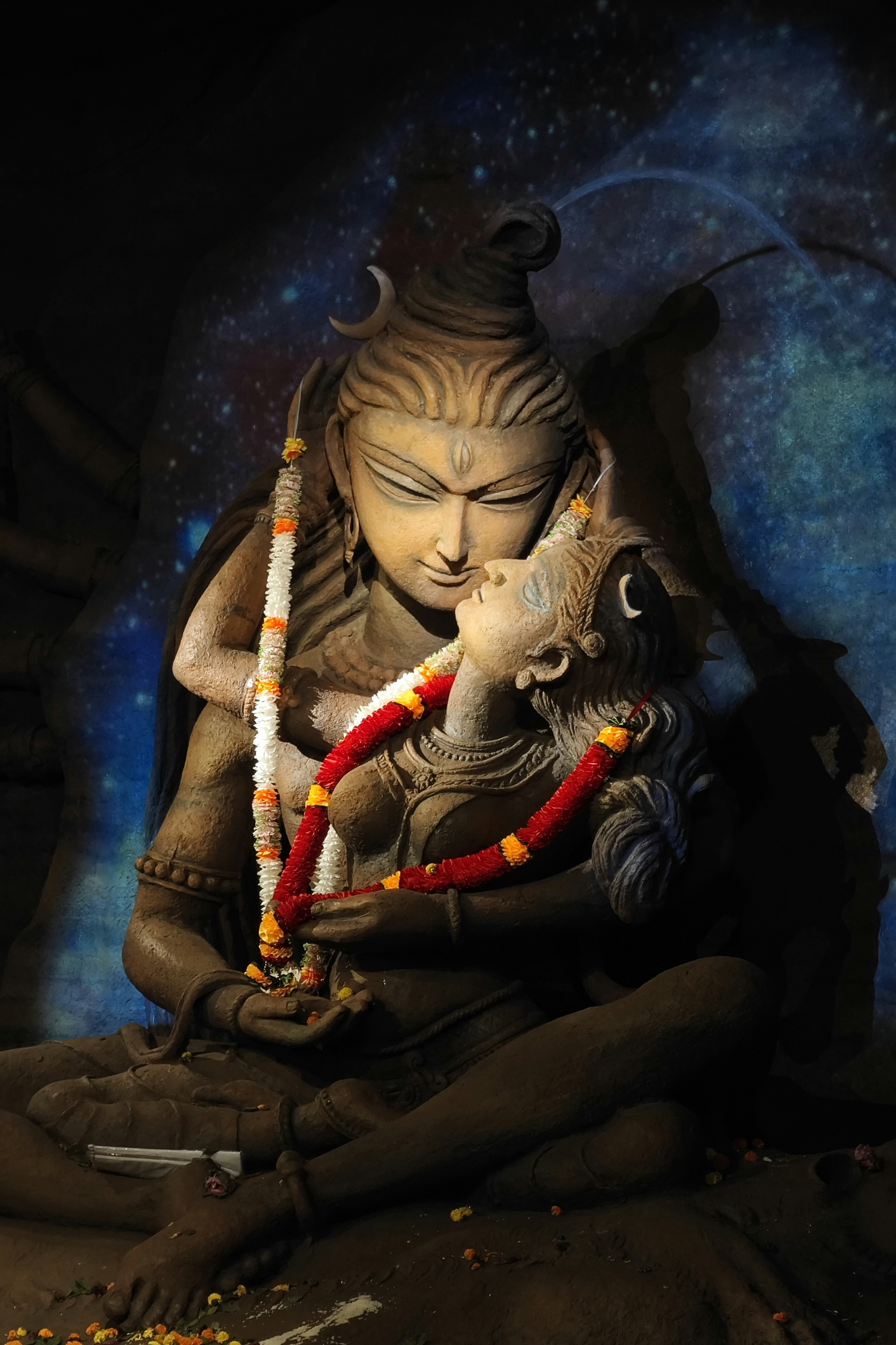 Stone statue of a seated couple embracing, adorned with flower garlands, set against a celestial mural background.