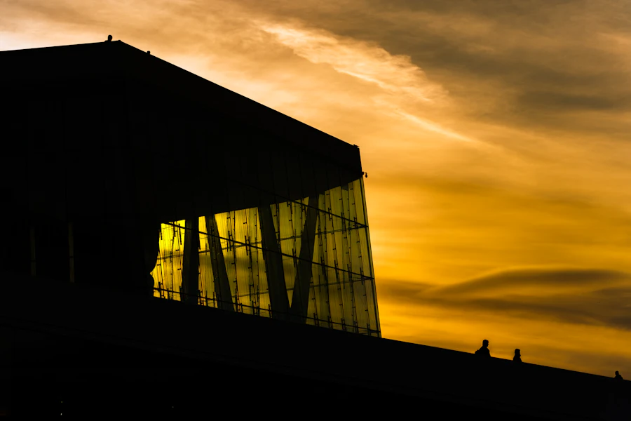 Dark angular building silhouette with a glowing glass section set against a dramatic golden sunset sky. Small figures stand along the rooftop edge.