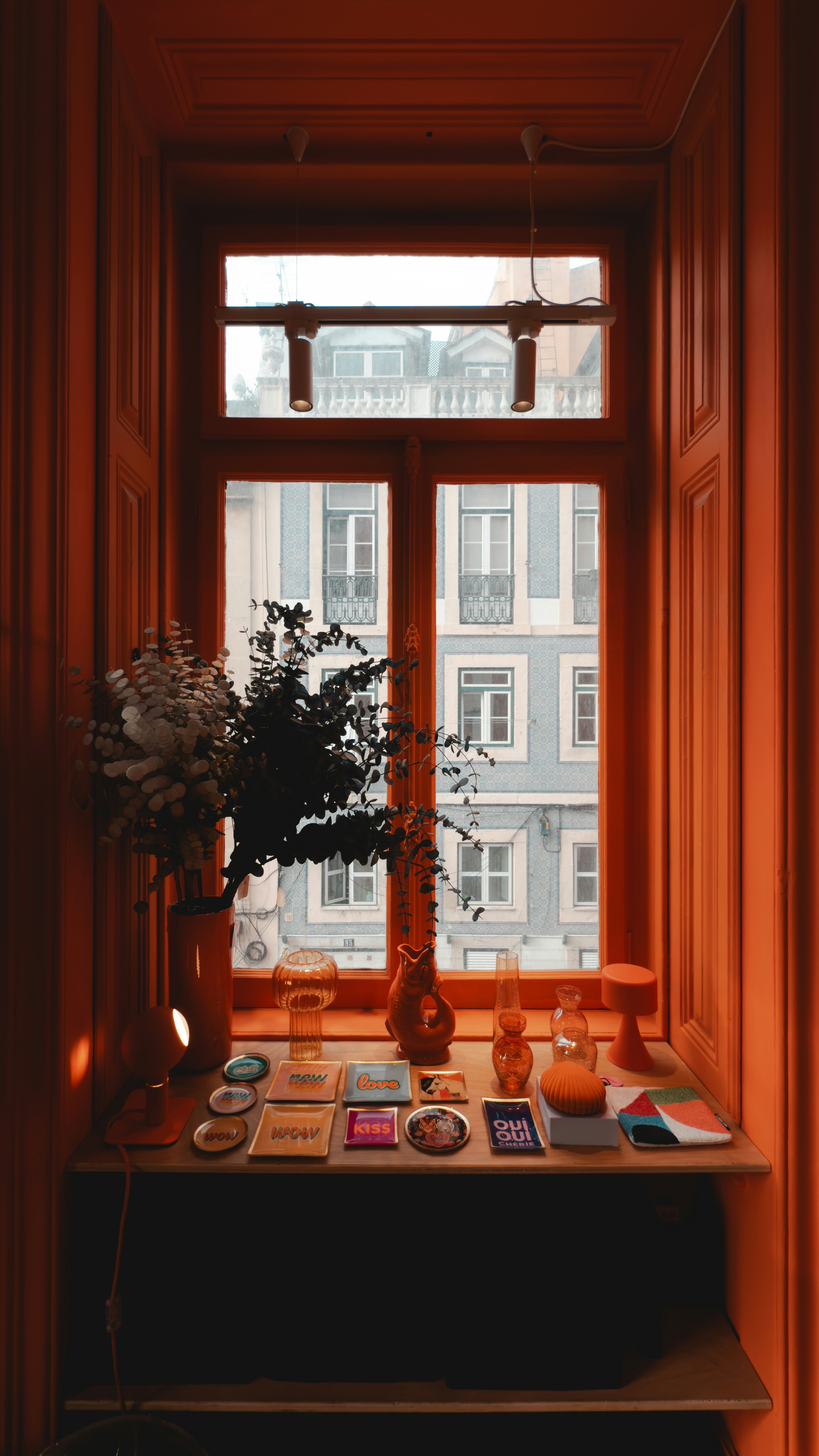 Warm-toned interior with a windowsill arrangement of plants, glassware, and knickknacks bathed in orange light. The view outside shows a city building through the window.