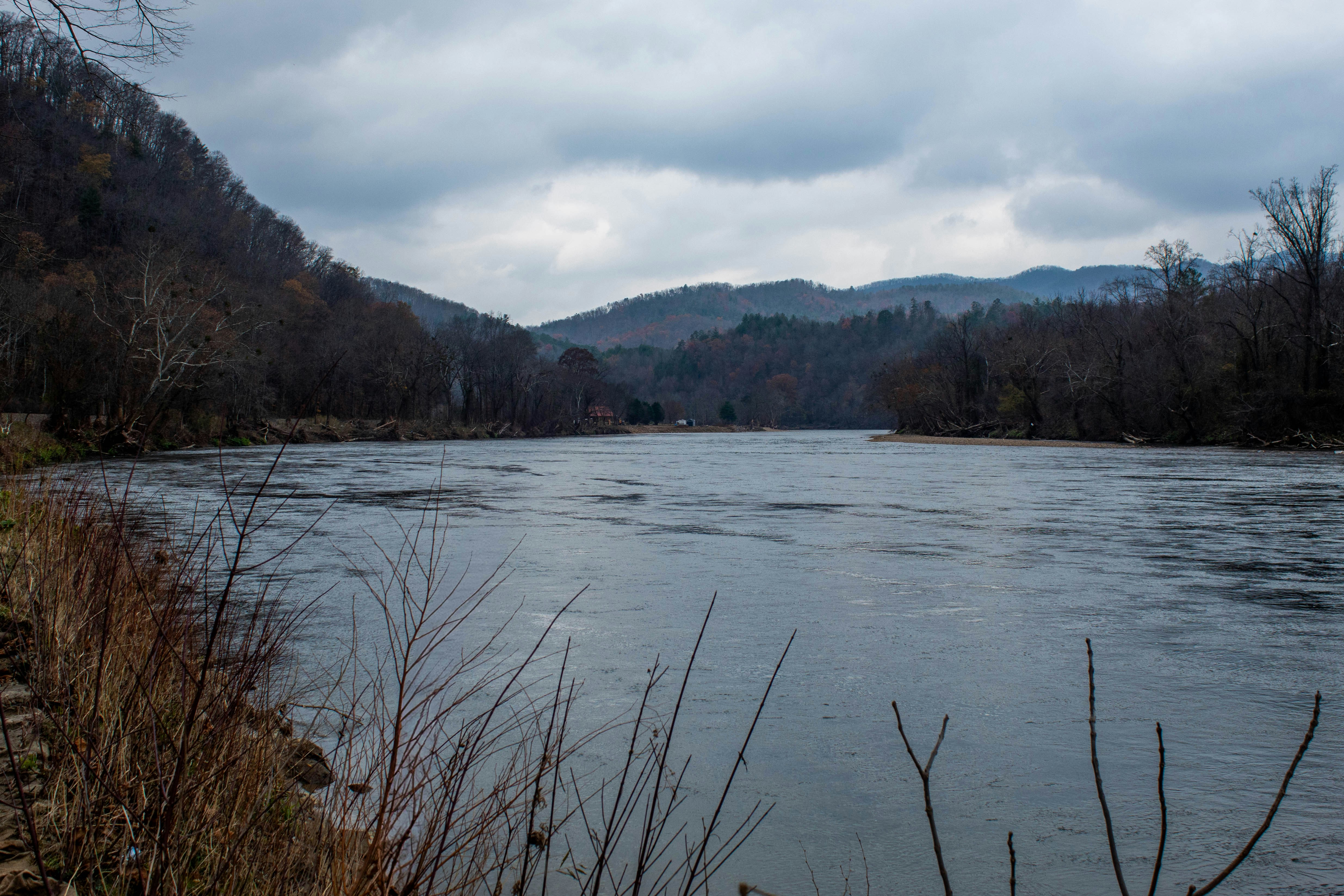 Calm river flowing through leafless trees with distant hills under a cloudy sky.