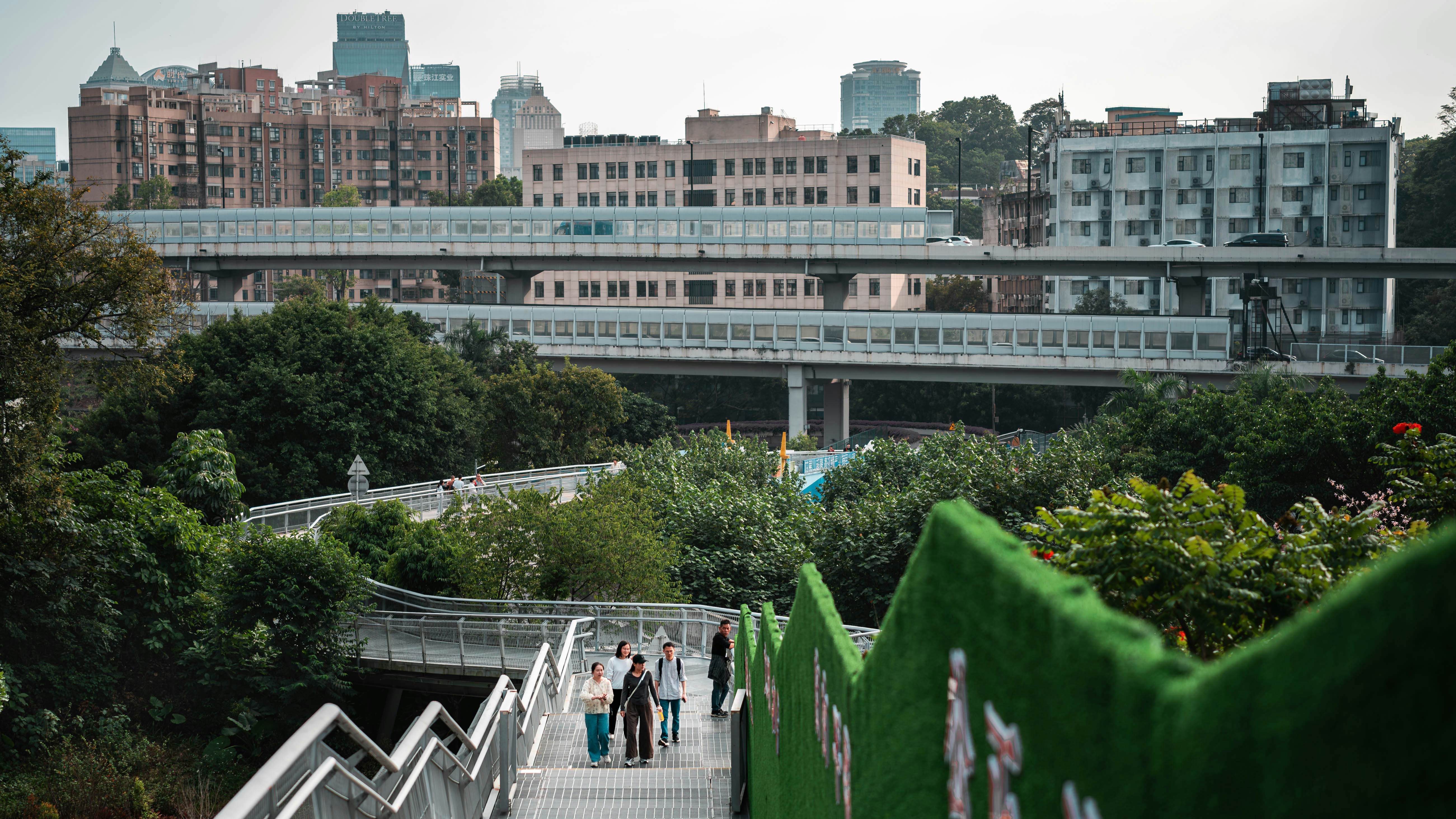 People walking on a staircase surrounded by green foliage with urban buildings and a bridge in the background.