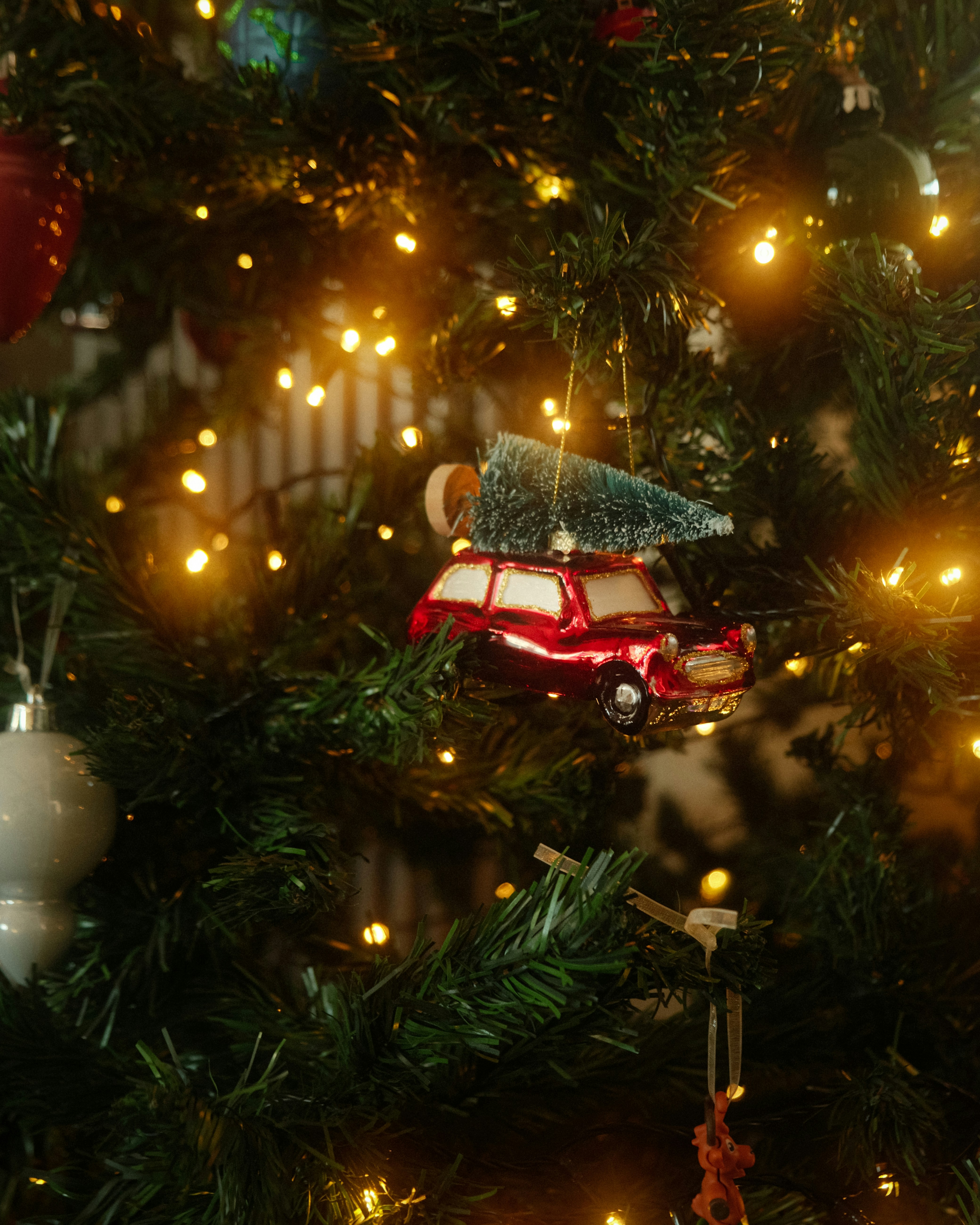 A tiny red toy car ornament hangs among warm fairy lights on a Christmas tree, a photograph capturing frosted details and pine needles framing the scene.