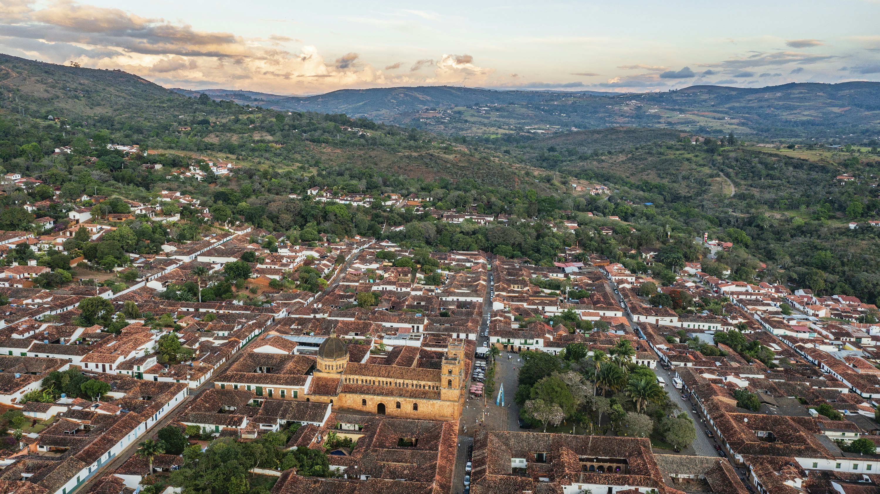 Aerial view of a historic town with red-tiled roofs nestled in lush green hills under a soft, pastel sky.