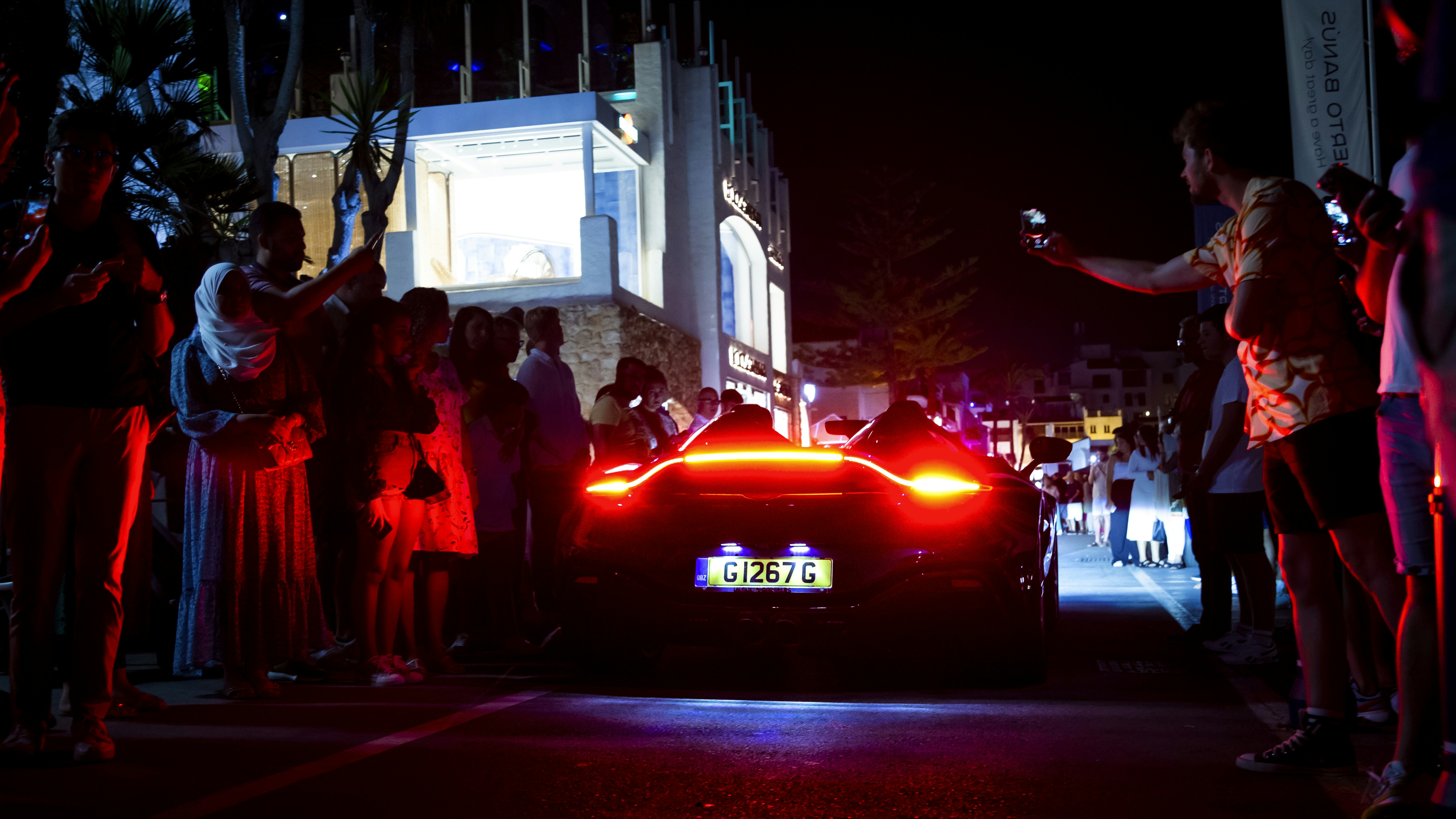 Group of people surrounding a brightly lit sports car on an urban street at night.