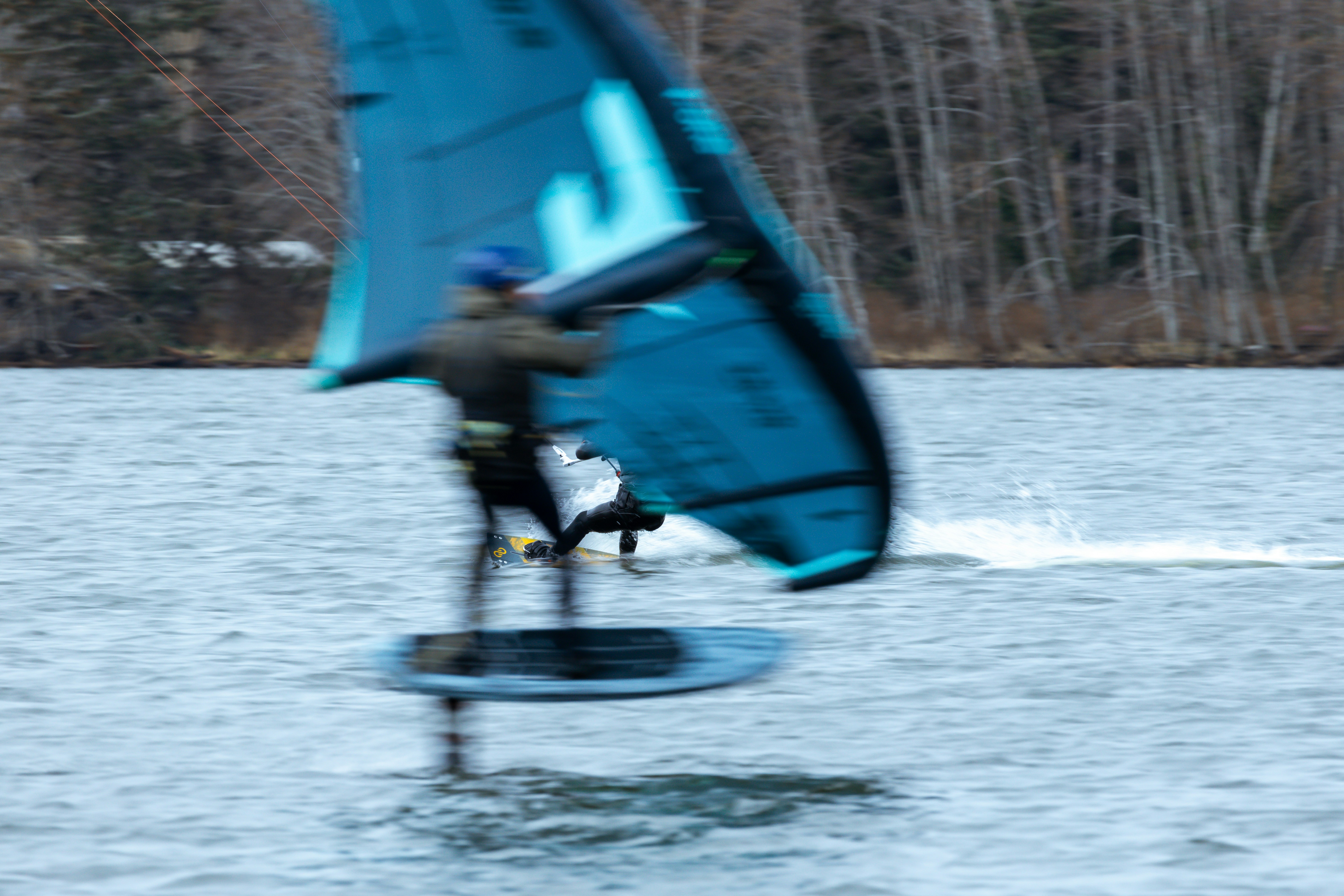 kite surfer eclipsed by a foil surfer on water | A person windsurfing on a lake with trees in the background