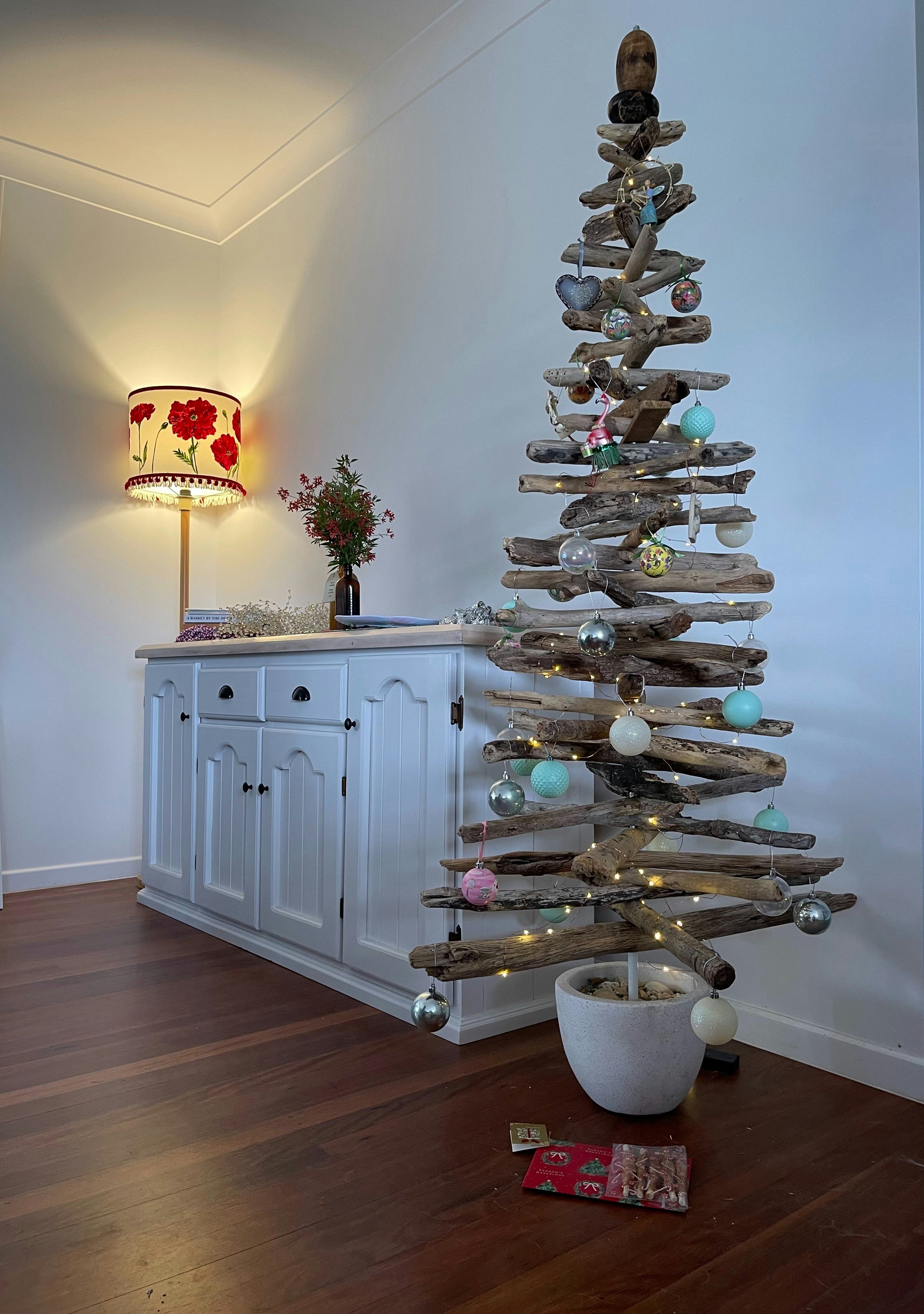 A creatively decorated driftwood Christmas tree adorned with pastel ornaments and fairy lights, positioned next to a vibrant floral lamp and a stylish sideboard.