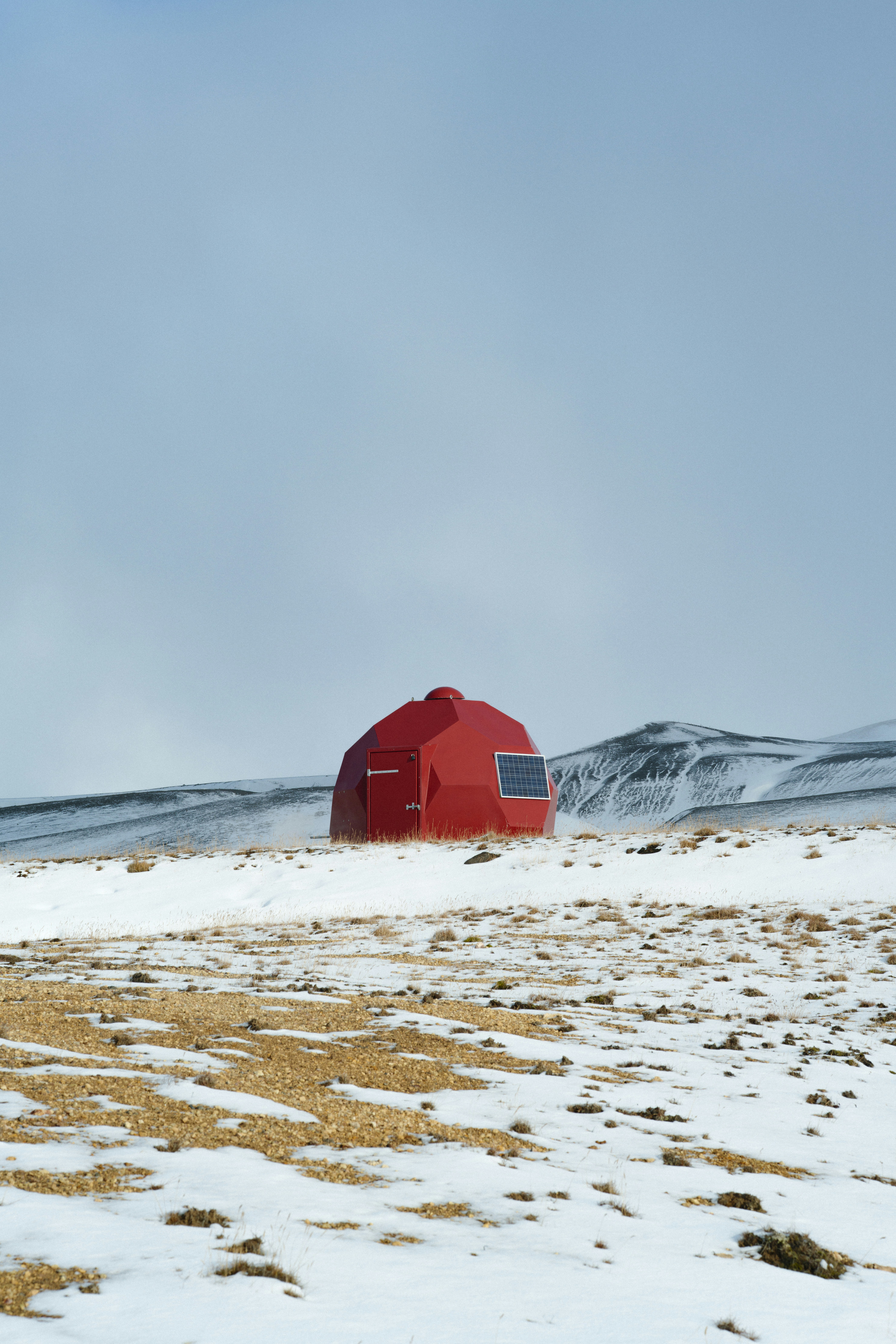 Red geodesic shelter with solar panel in snowy landscape against overcast sky.