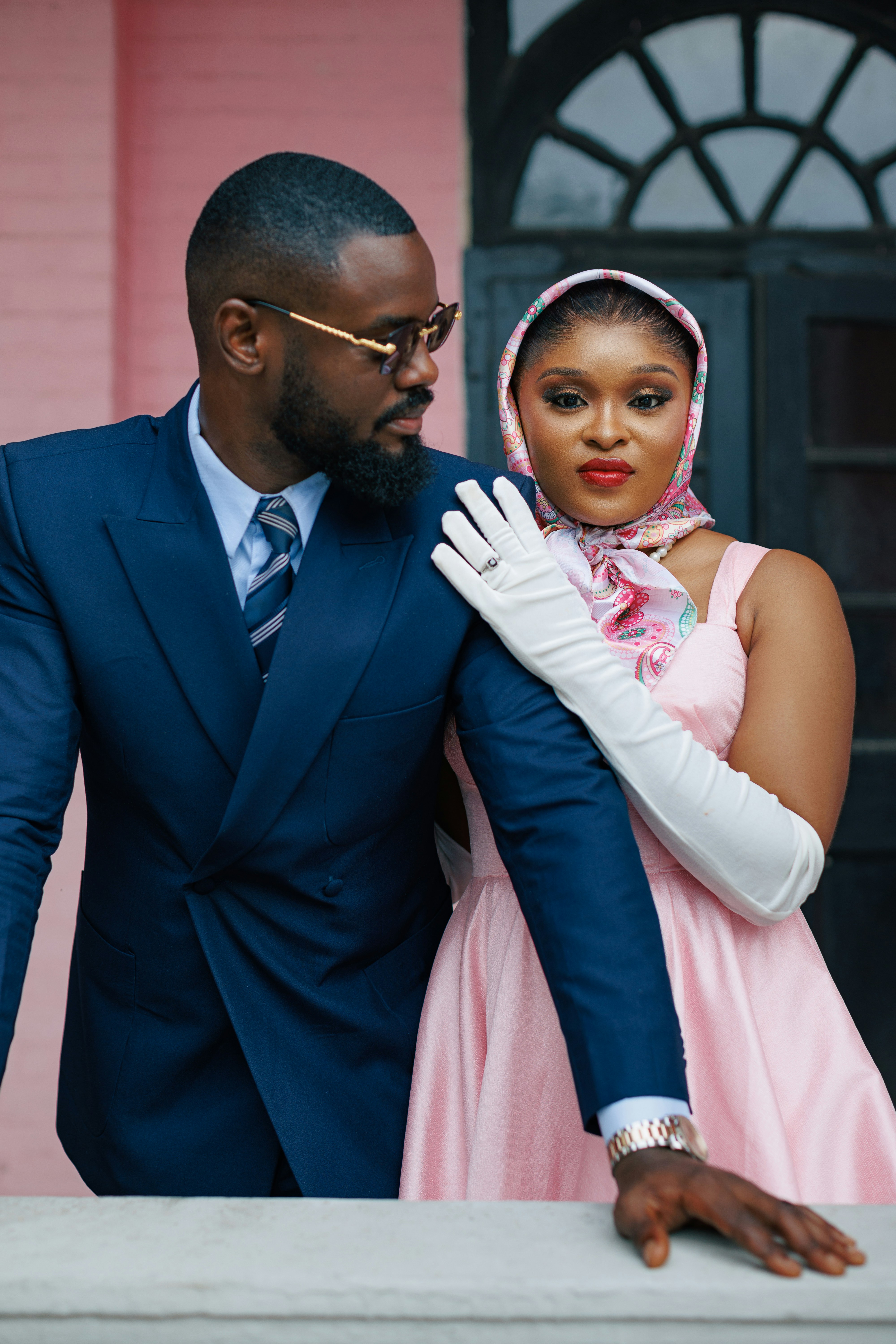 Sophisticated couple in navy suit and pink dress poses against a soft pink backdrop.