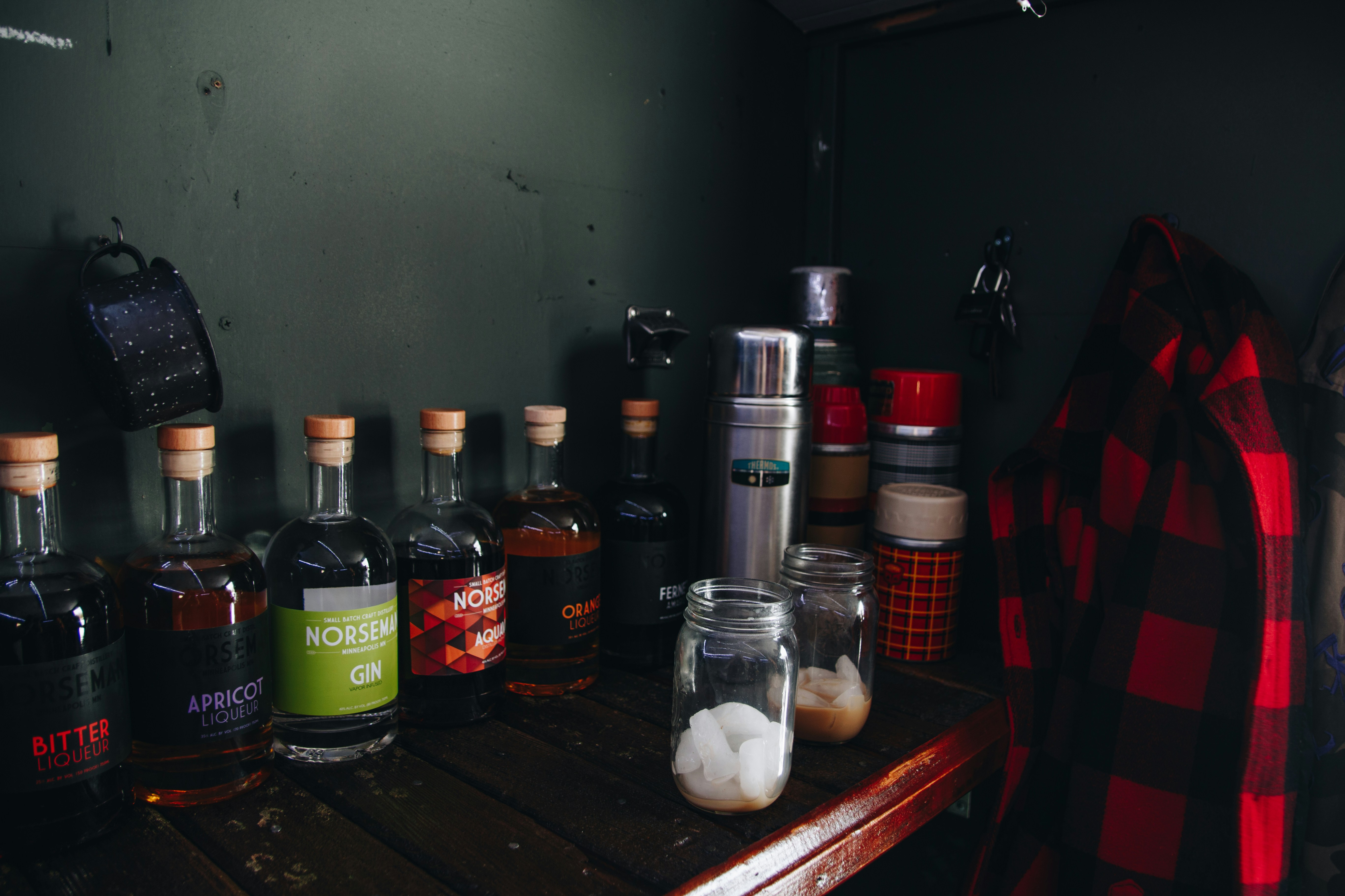 Whispered Spirits on a Dim Bar Shelf: Bottles, Sugar Jar, and a Plaid Coat