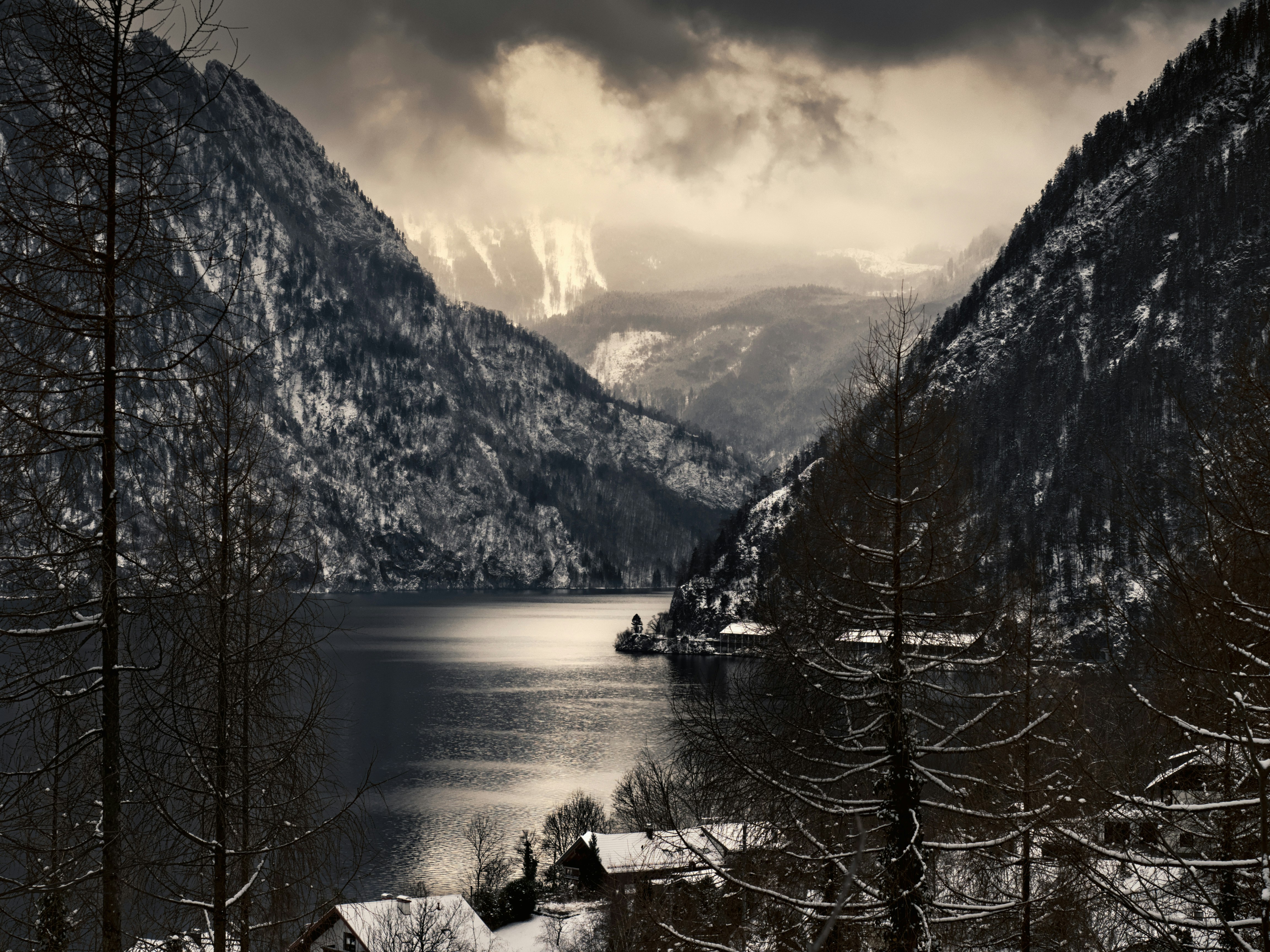 Snow-covered mountains encircle a tranquil lake under a moody sky, with a distant village nestled by the shore.