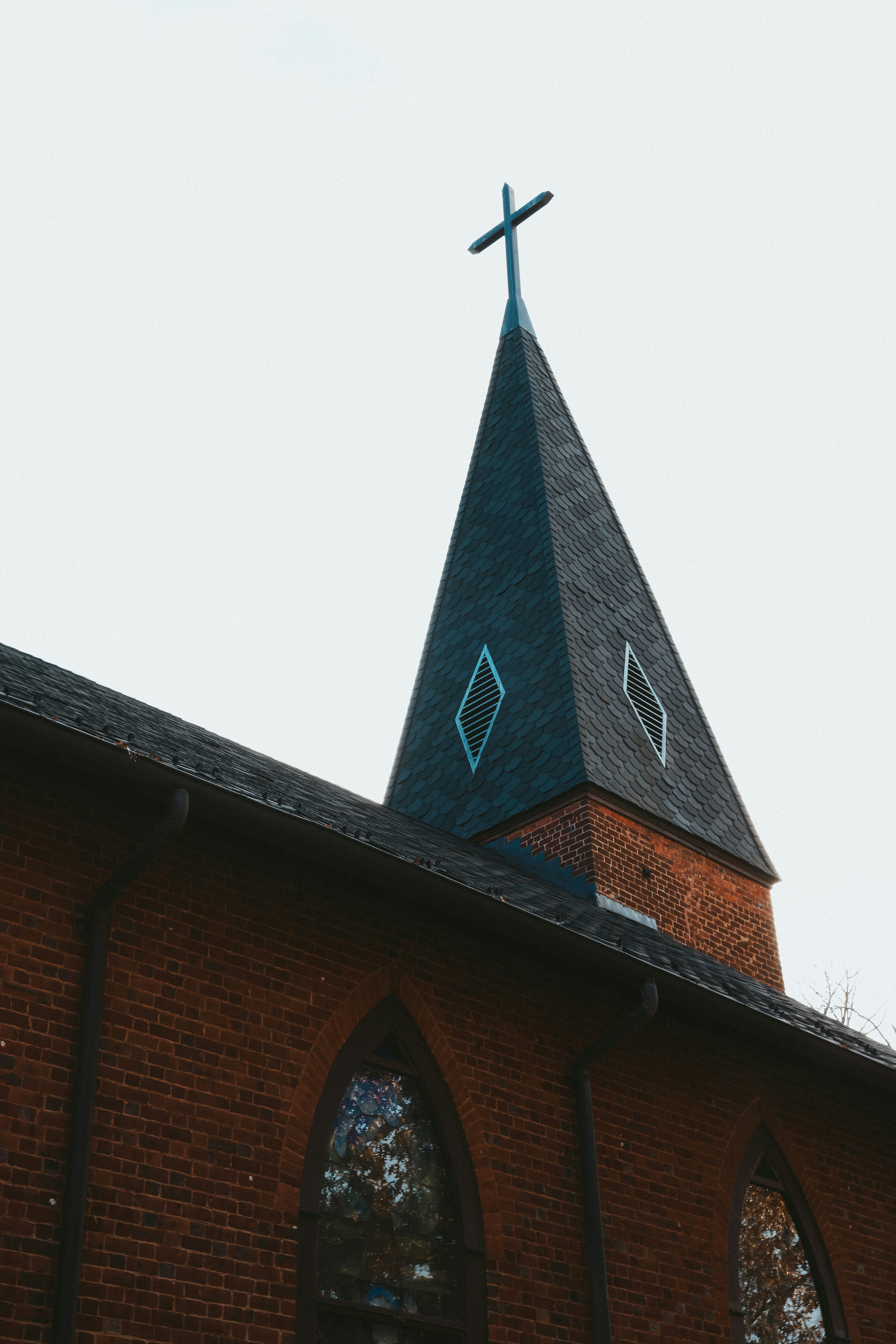 Brick church façade with a tall slate steeple topped by a cross rises against a pale, overcast sky. Arched windows along the facade add vertical rhythm.