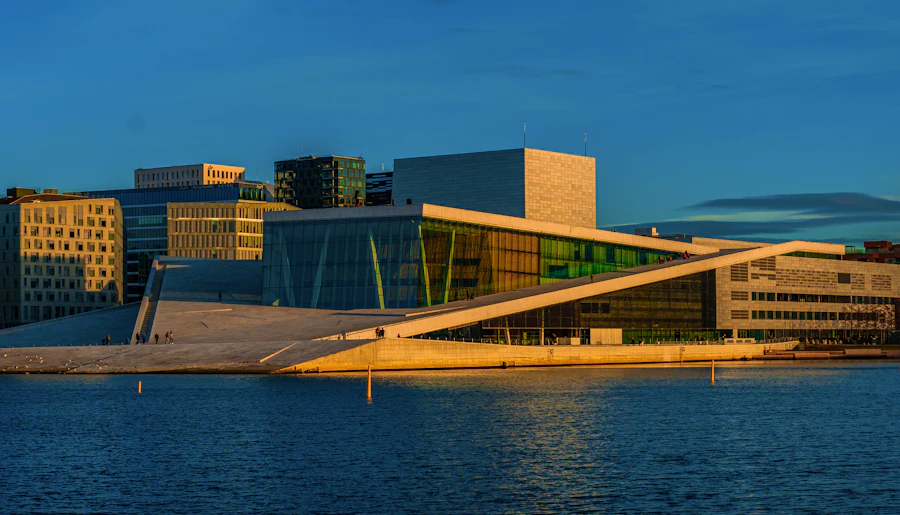 Modern angular waterfront building complex glowing in warm golden light across calm blue water at dusk. Clear sky and reflections emphasize the architecture's sharp lines.