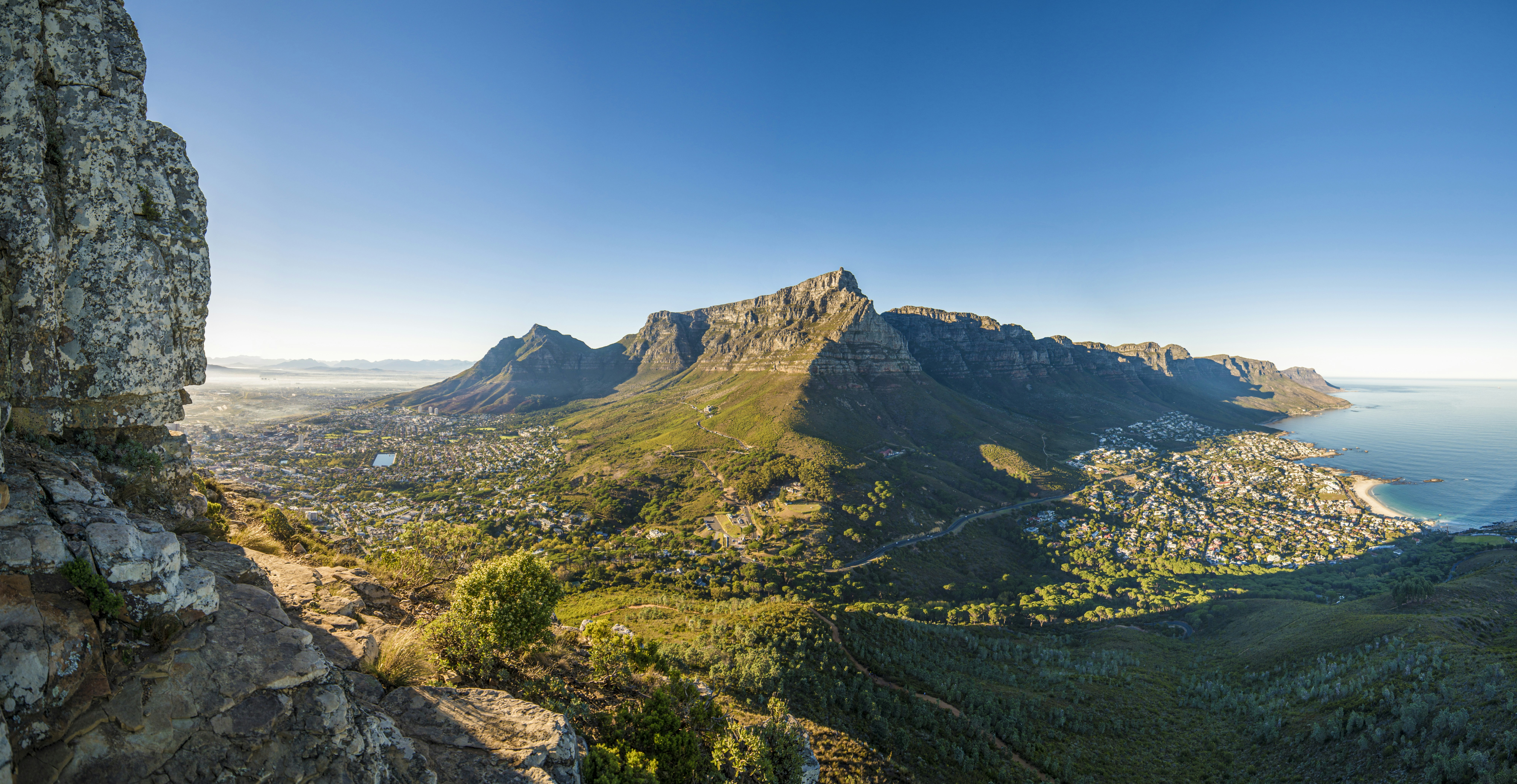 Panoramic view of a sunlit mountain range with lush greenery and a distant ocean under a clear blue sky.