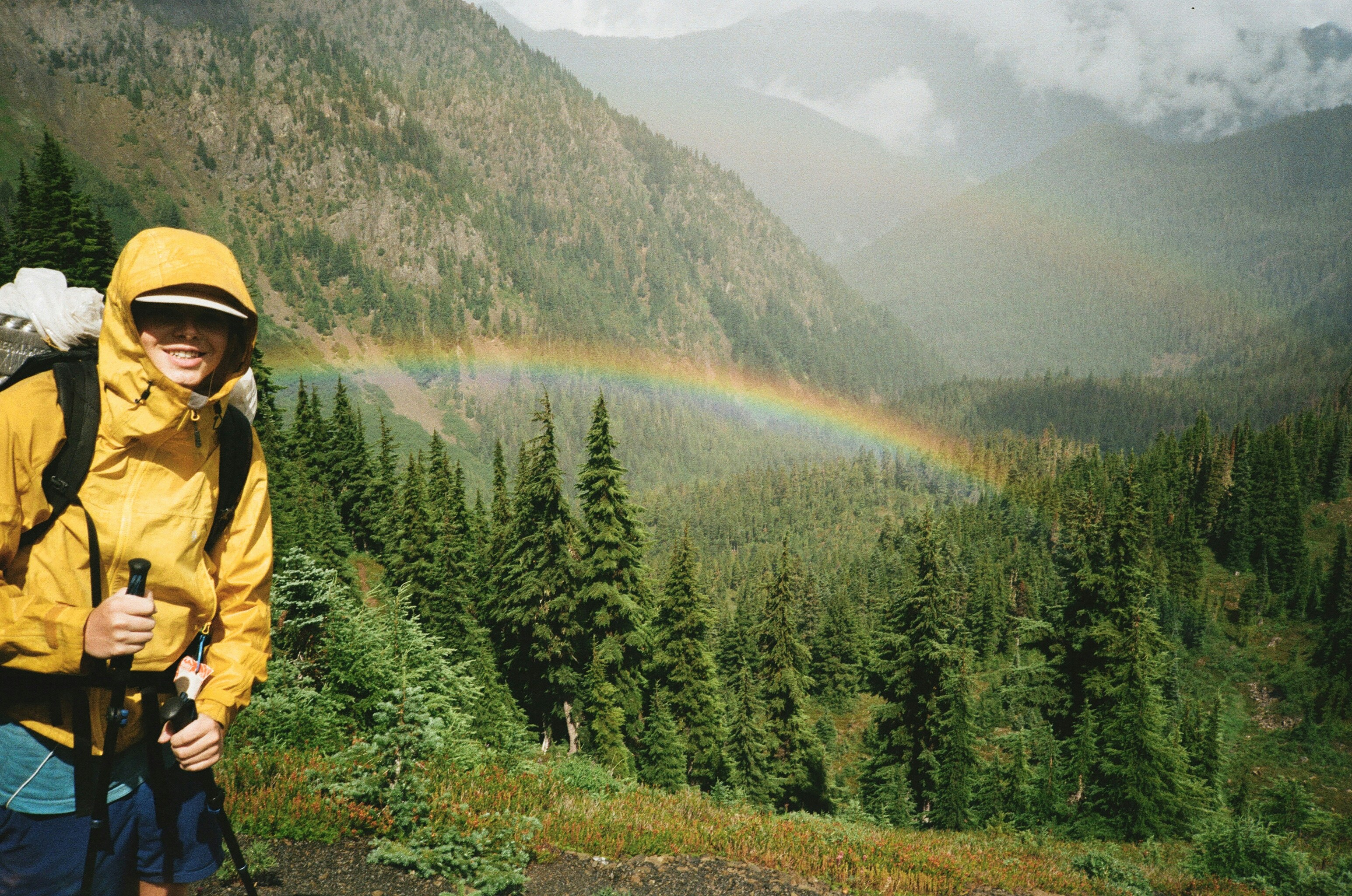 A hiker in a bright yellow rain jacket stands on a trail amid a dense conifer forest, with a vivid rainbow arching across the valley toward rugged mountains.
