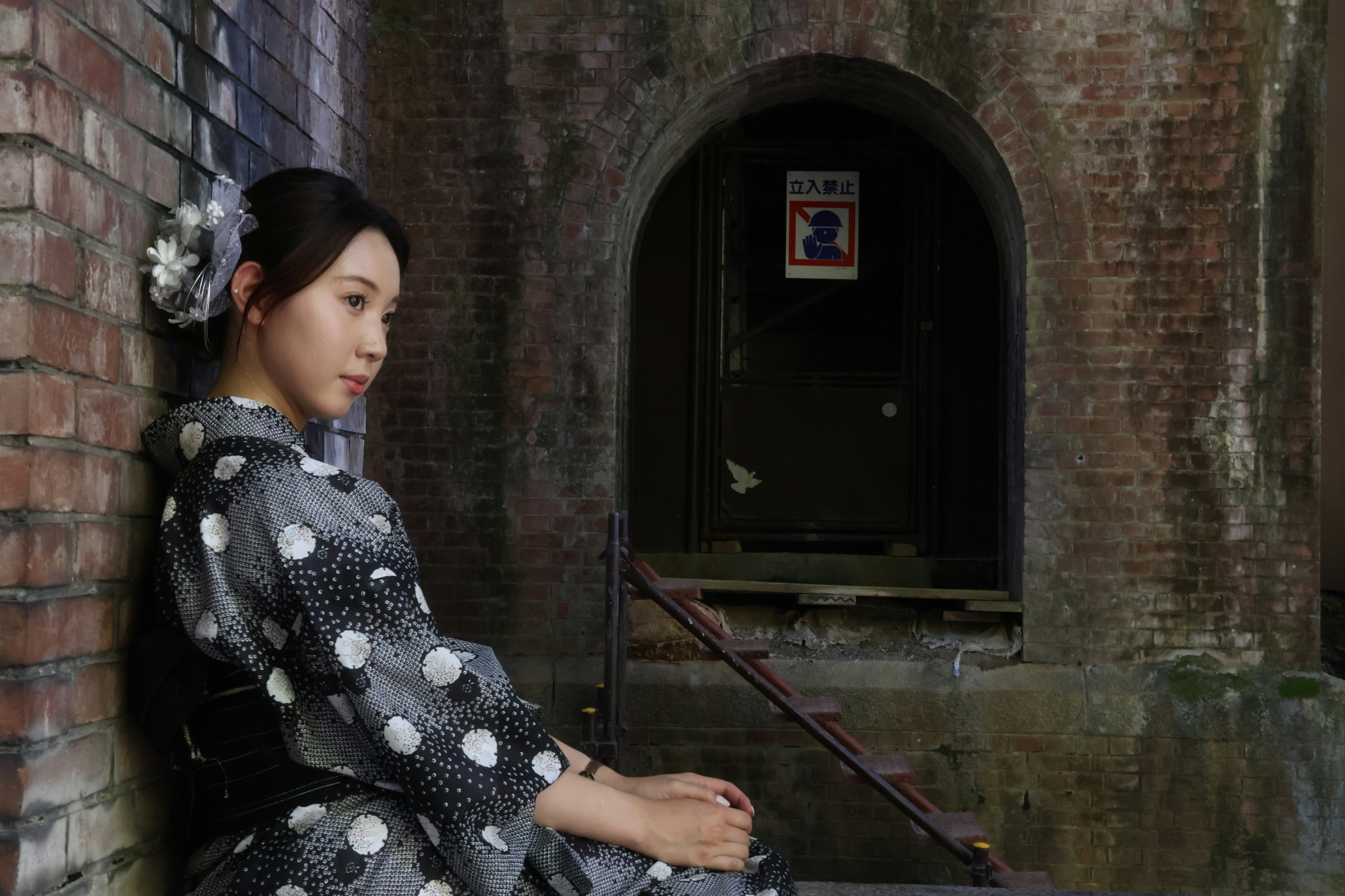 A woman in a traditional kimono sits gracefully against a textured brick wall, framed by an arched doorway. The scene captures a blend of cultural heritage and modern architecture.