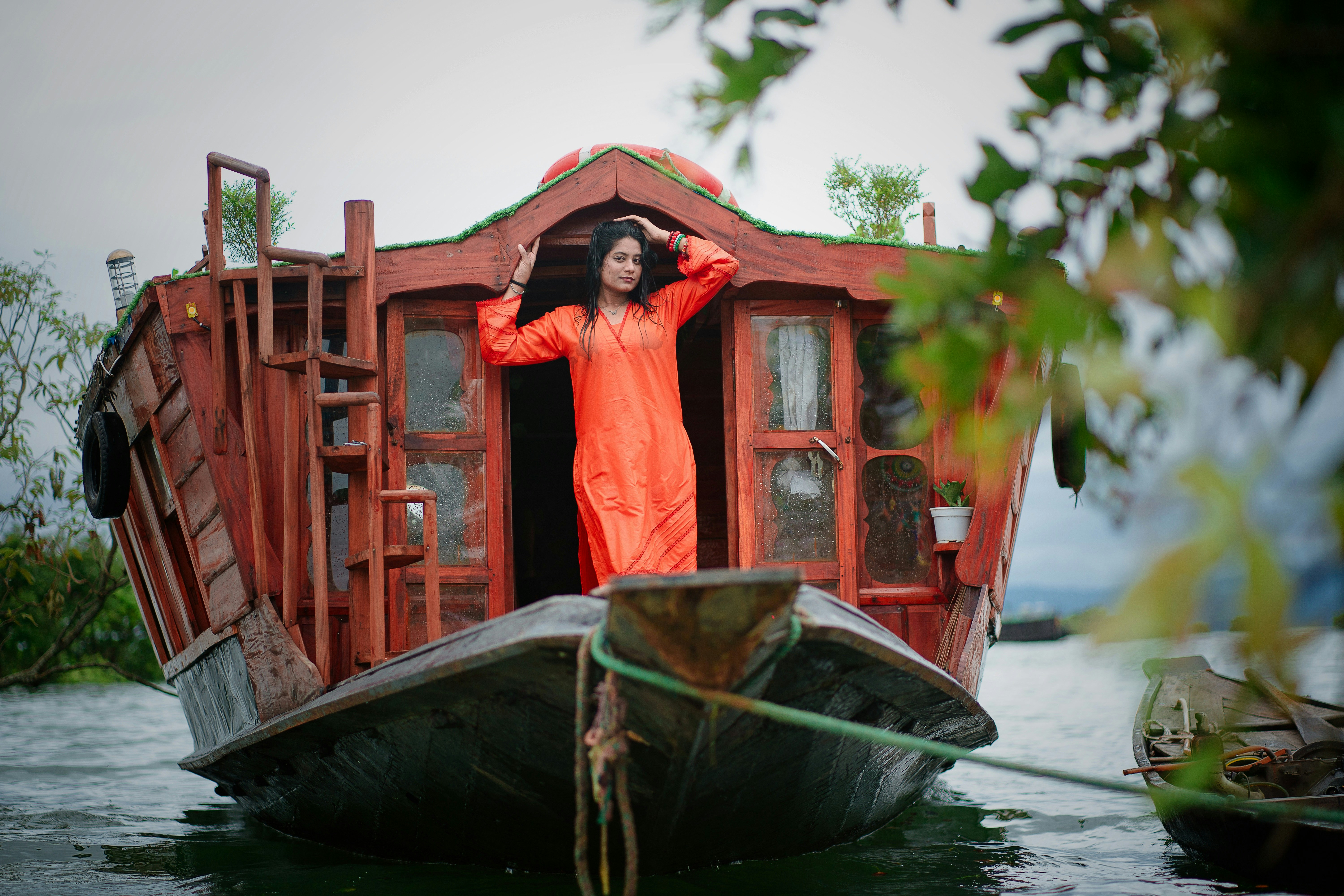 Person in an orange outfit stands on the deck of a rustic wooden boat surrounded by water and greenery.