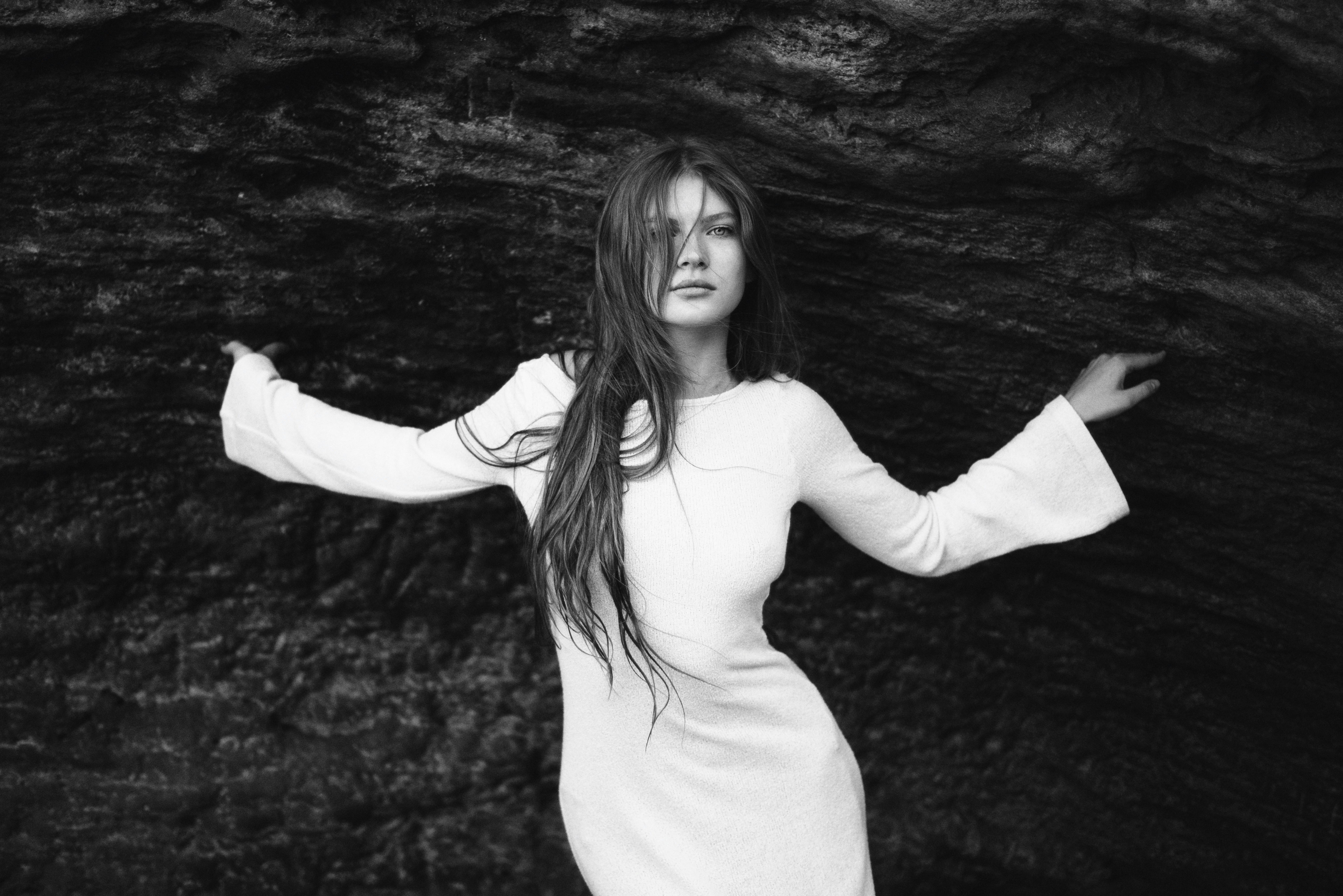 Woman in a flowing white dress stands against a textured rock backdrop, arms gracefully extended.
