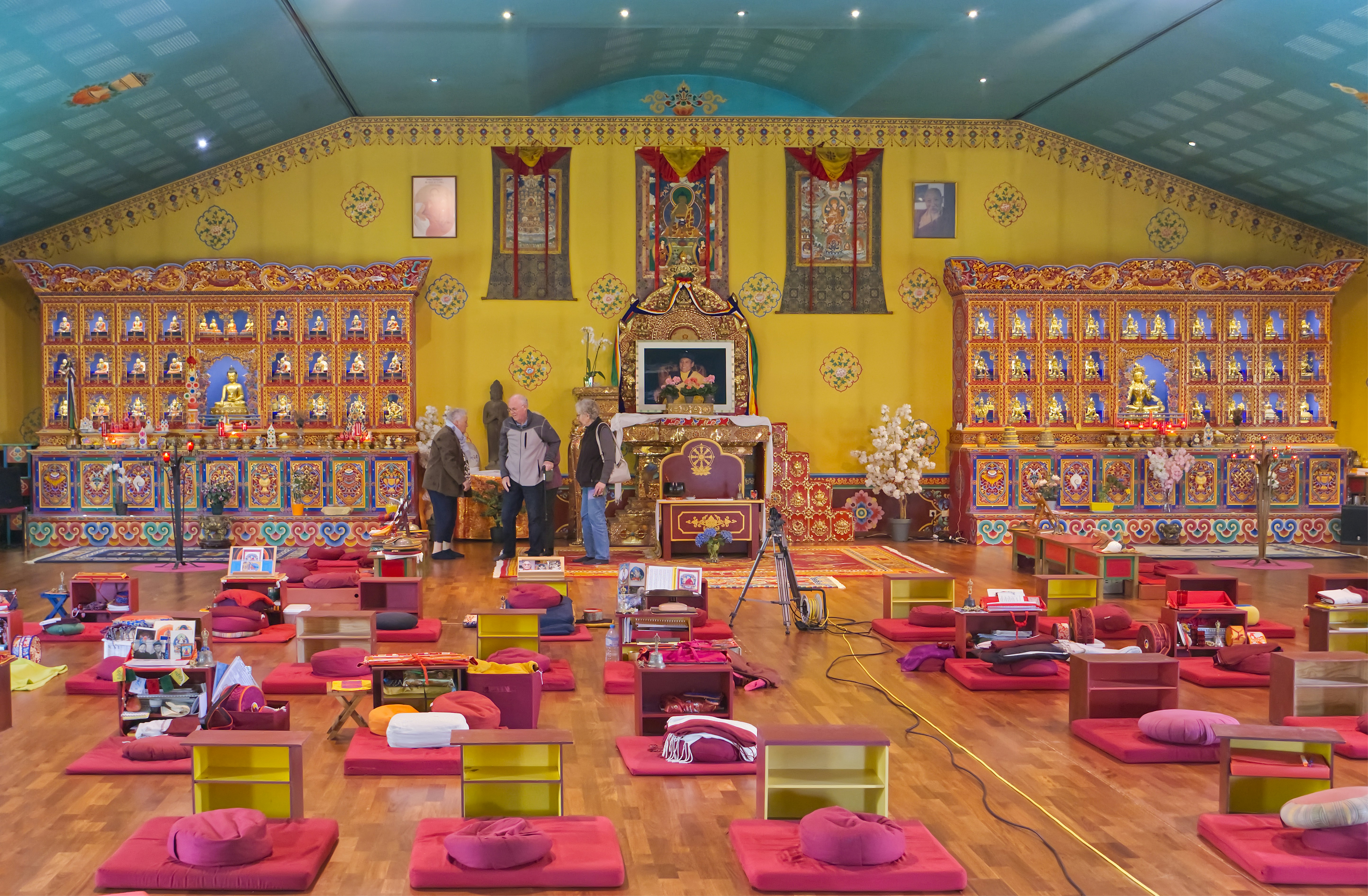 Cette photographie nous plonge dans le centre Drukpa Plouray dans le Morbihan. Ce lieu de culte bouddhiste est très coloré et richement décoré. Le décor est dominé par des couleurs chaudes comme le jaune, le rouge et l'orange, créant une atmosphère chaleureuse et accueillante. L'espace spirituel est structuré de manière symétrique, avec un autel central surmonté d'une statue en or représentant une divinité bouddhiste. Des tissus rouges ornés de motifs décoratifs encadrent l'autel, conférant une dimension sacrée à cet espace. | A large room filled with lots of colorful furniture