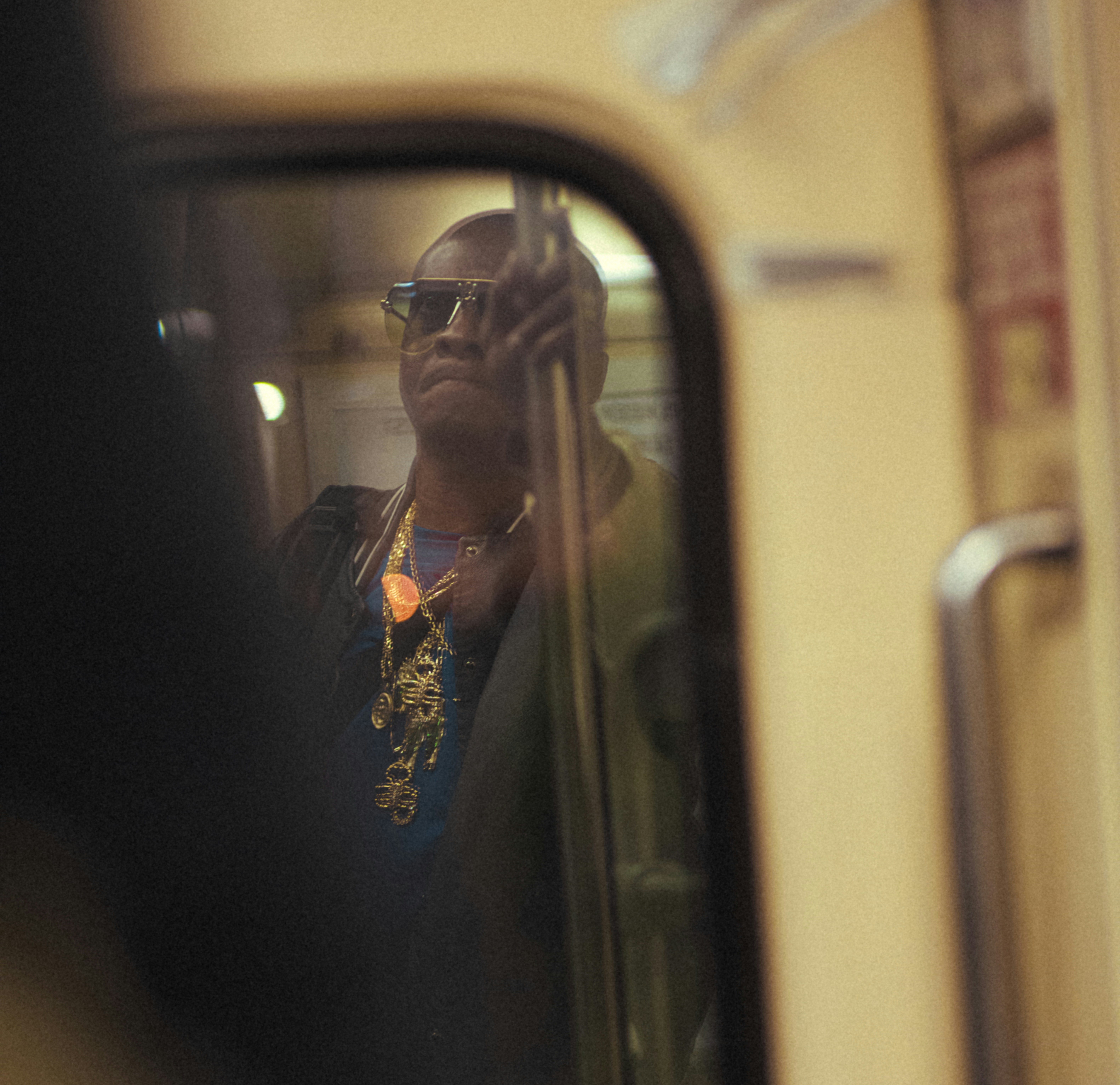 A photograph of a man wearing sunglasses and a gold necklace, reflected in a vehicle window with warm amber lighting.