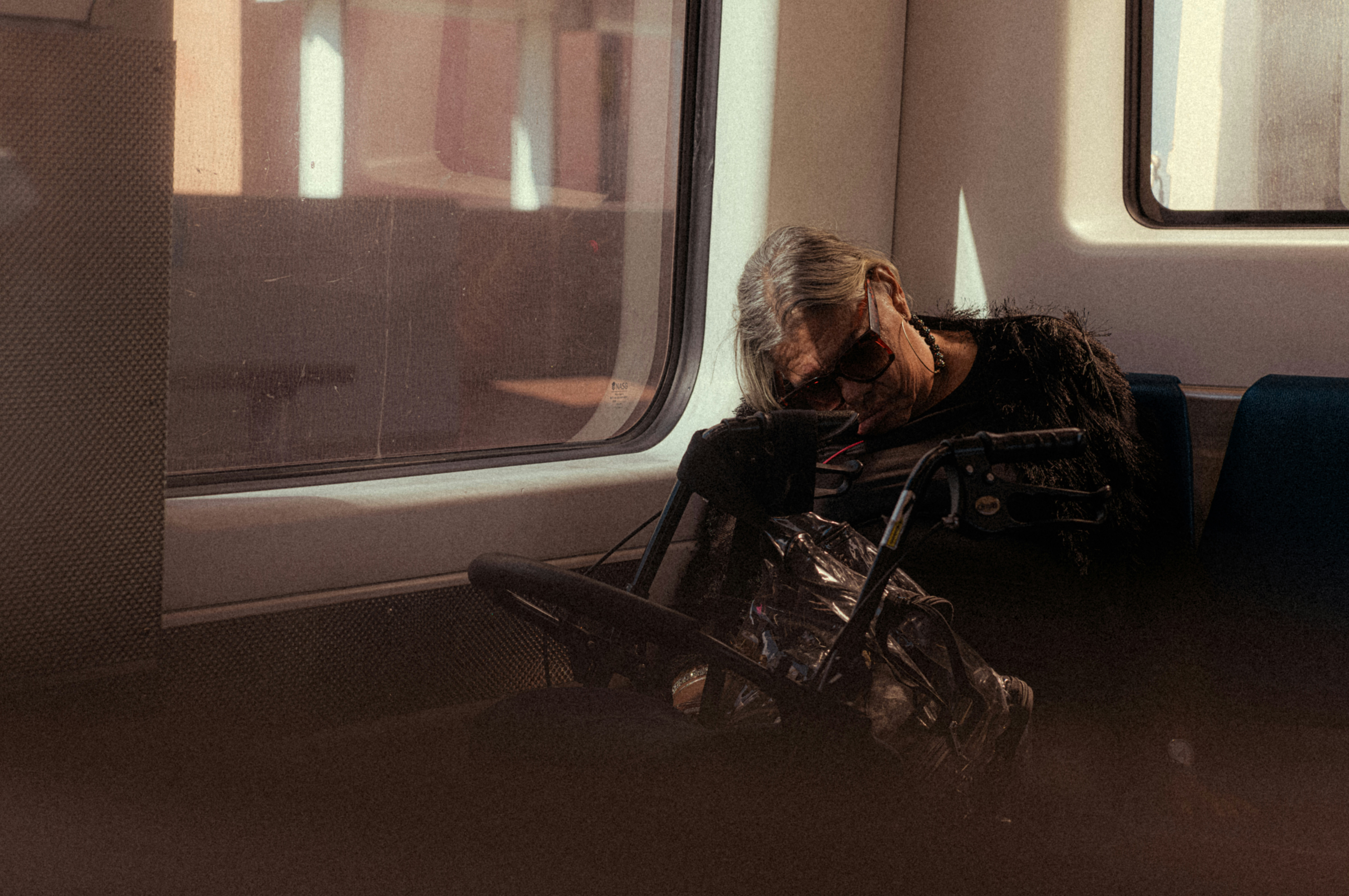 Woman on a train, head resting by a device or prop, soft warm light through window; a portrait-like moment with a hint of travel mood. The scene appears to be an indoor setting, possibly a conveyance interior.