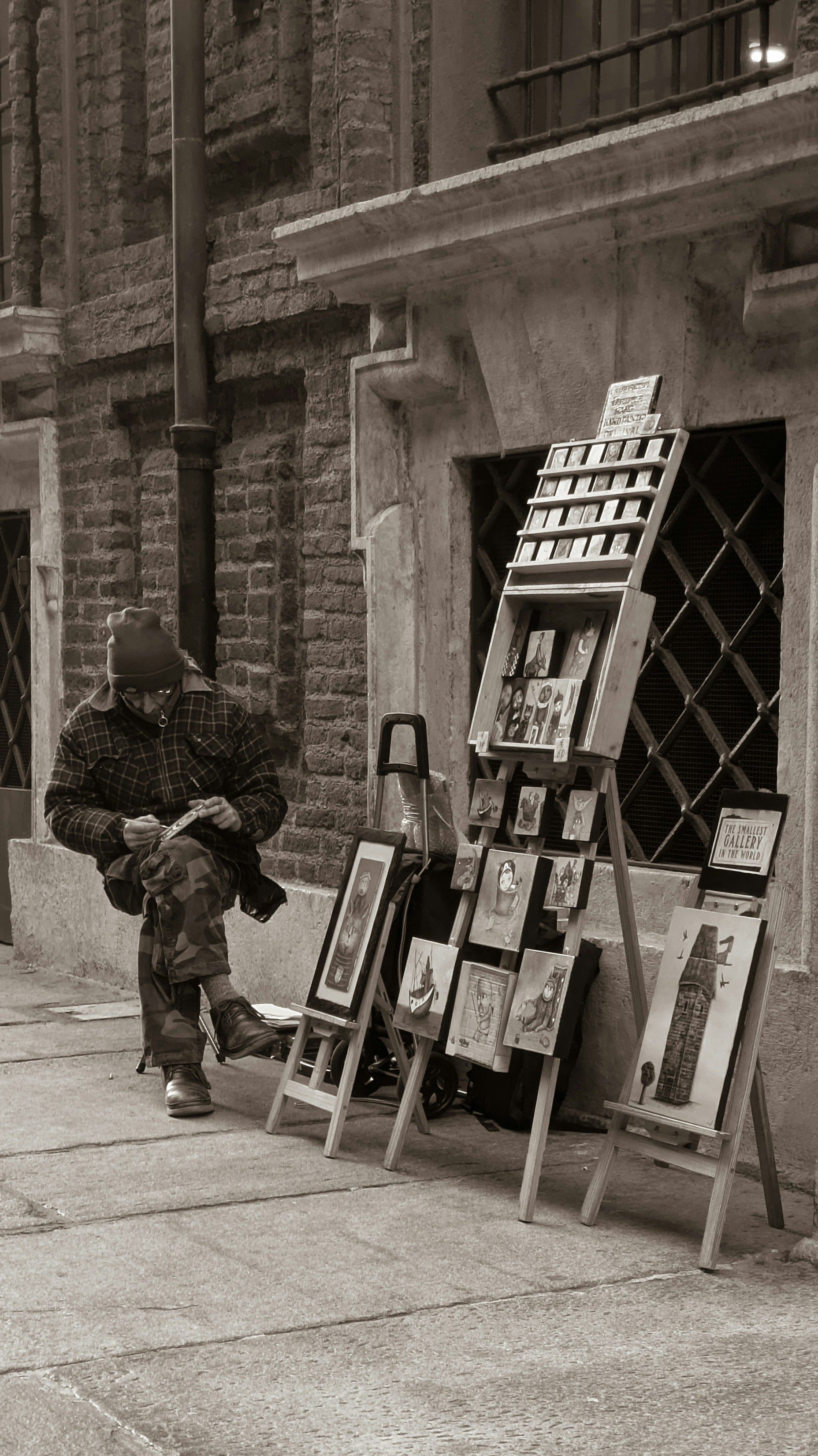 Artist arranging paintings on a cobblestone street beside a brick building, captured in sepia tones.