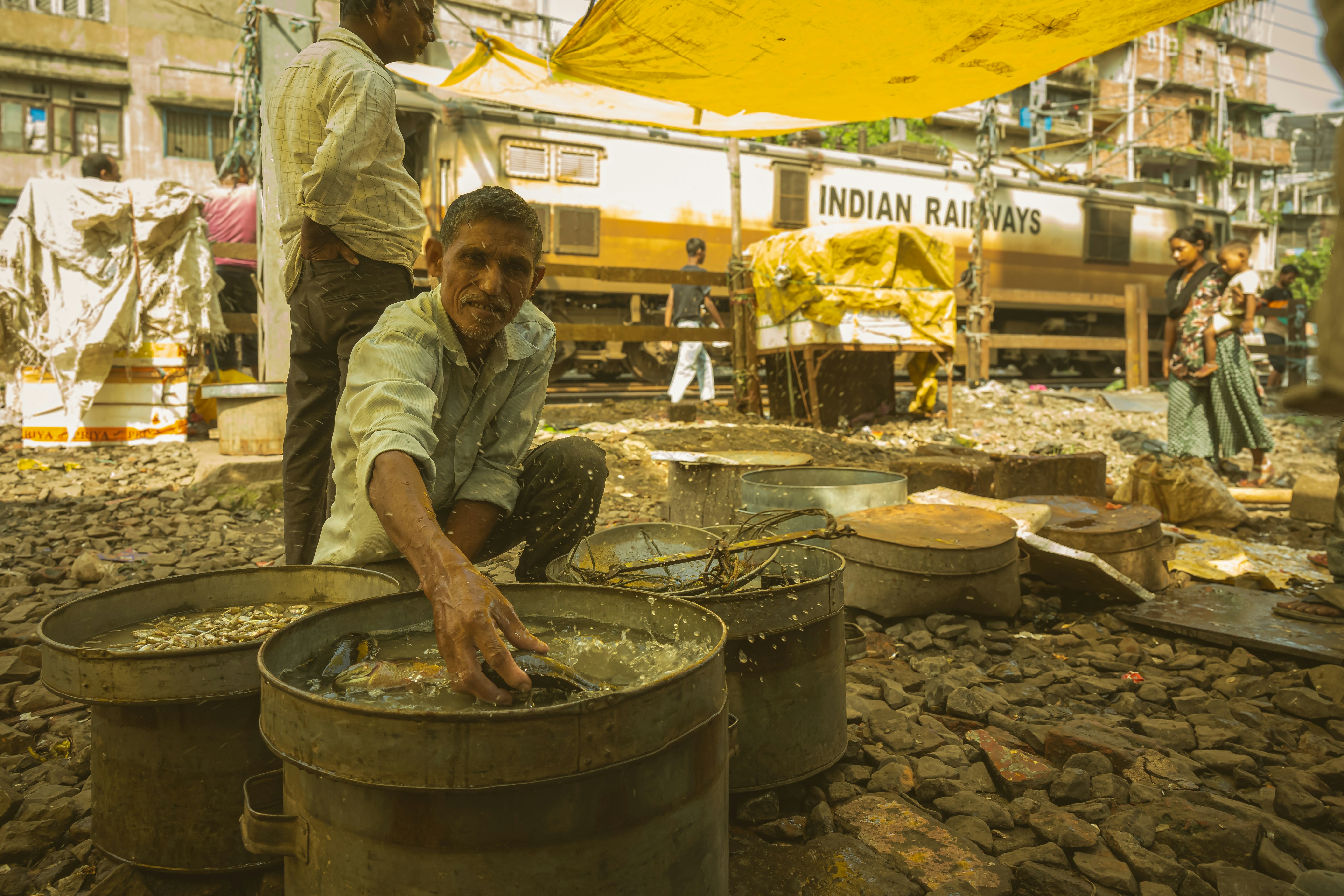 Vendor arranging fish in metal tubs under a yellow canopy at an outdoor market.