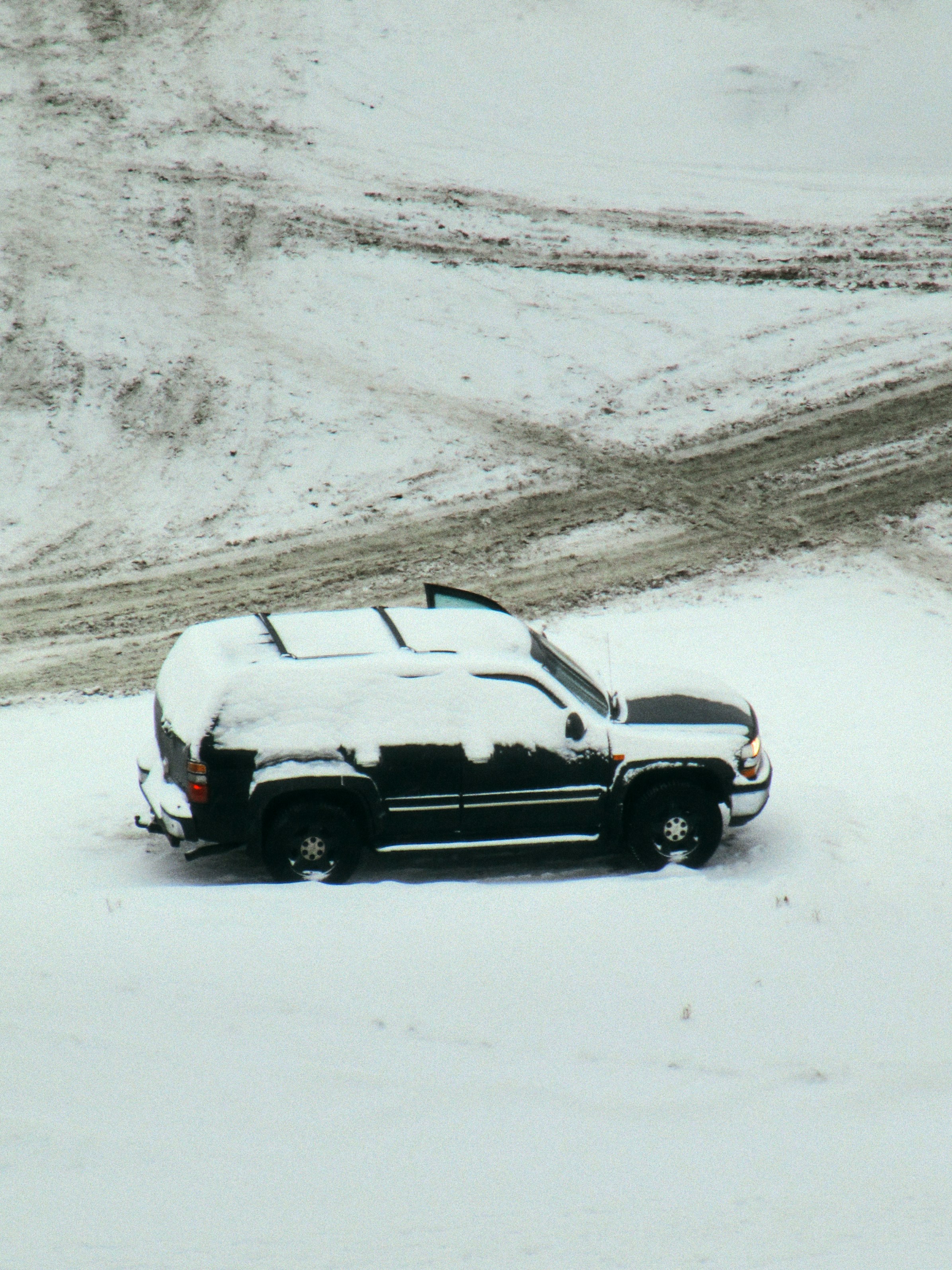 Black pickup truck sits half-buried in fresh snow, its silhouette stark against a quiet, wintry landscape. Snow-dusted hills and faint tire tracks frame the vehicle.