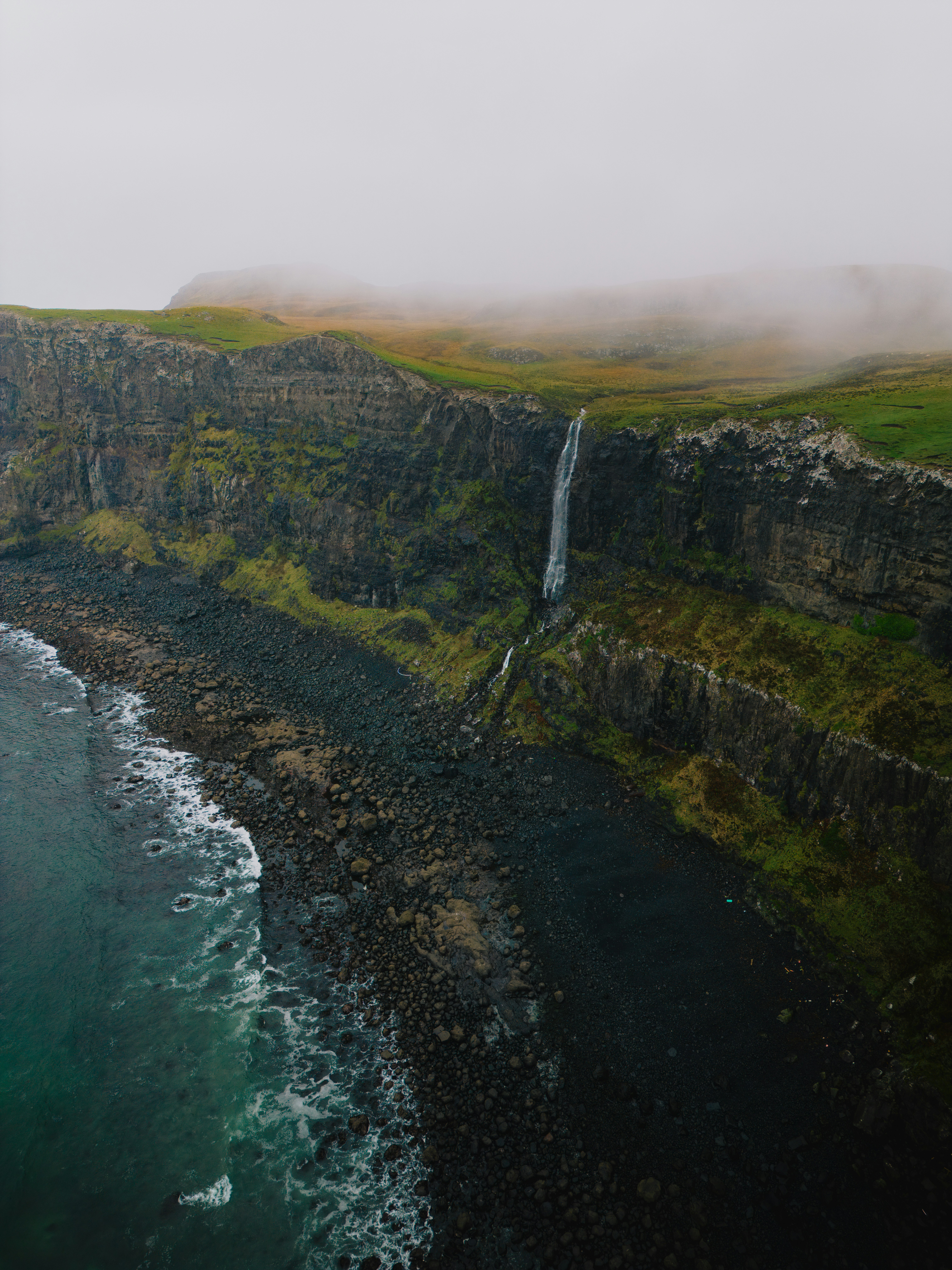 Waterfall cascading down a misty cliffside into a rocky shoreline, with overcast skies above.