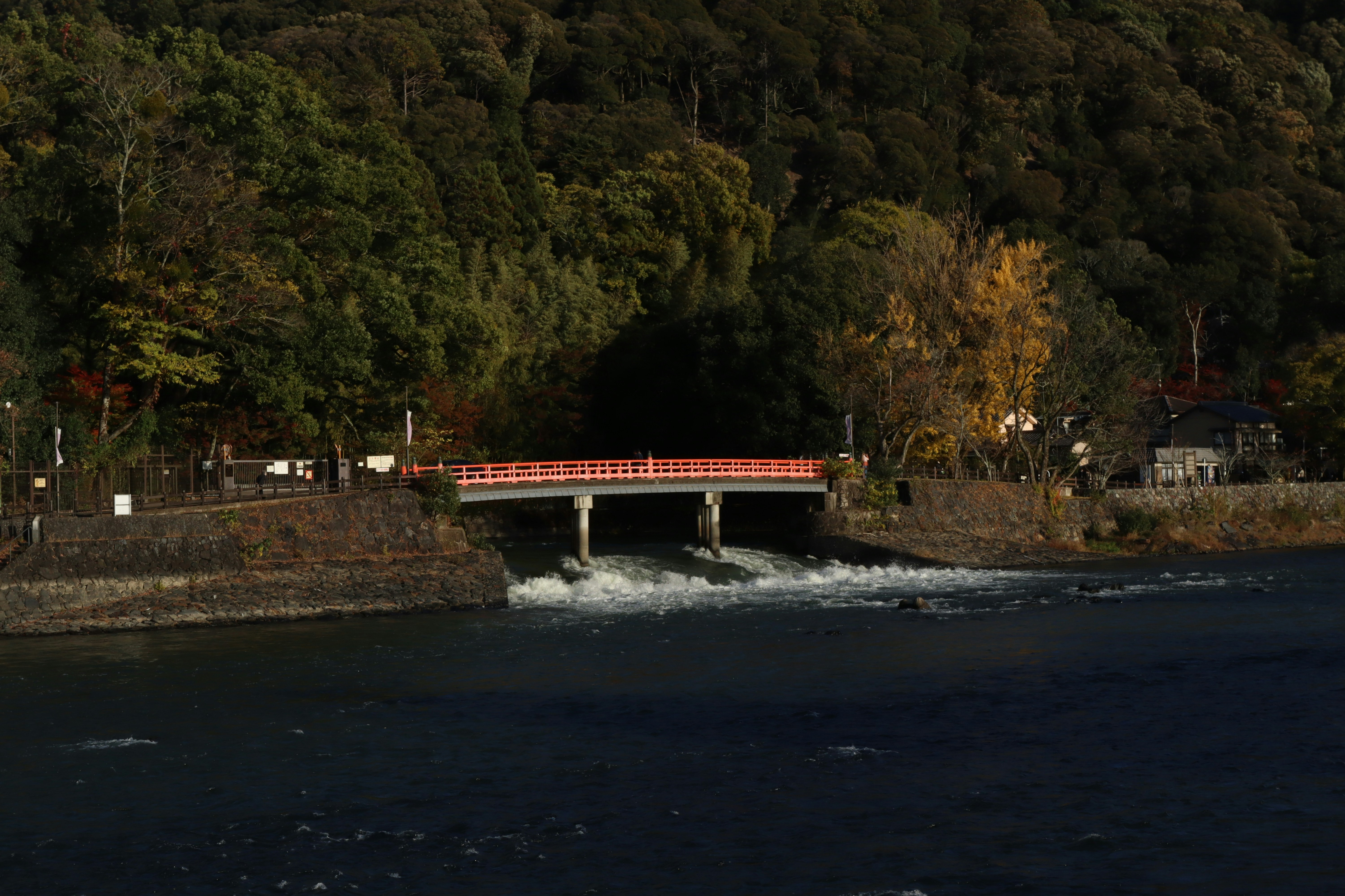 A red bridge spans a river with gentle rapids, framed by trees with autumn foliage.