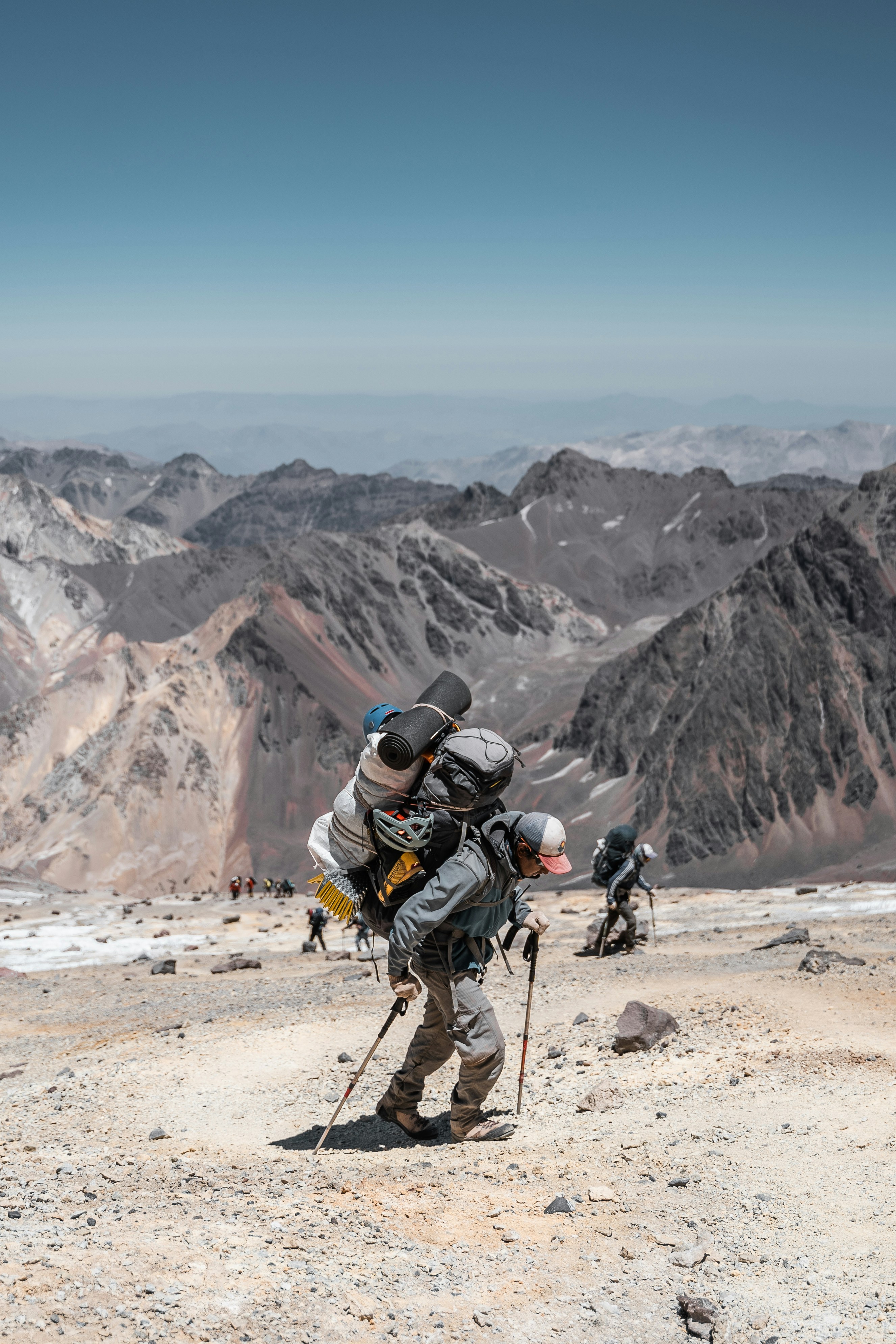 Hikers ascending a rugged mountain range under a clear blue sky, with expansive rocky terrain in the background.