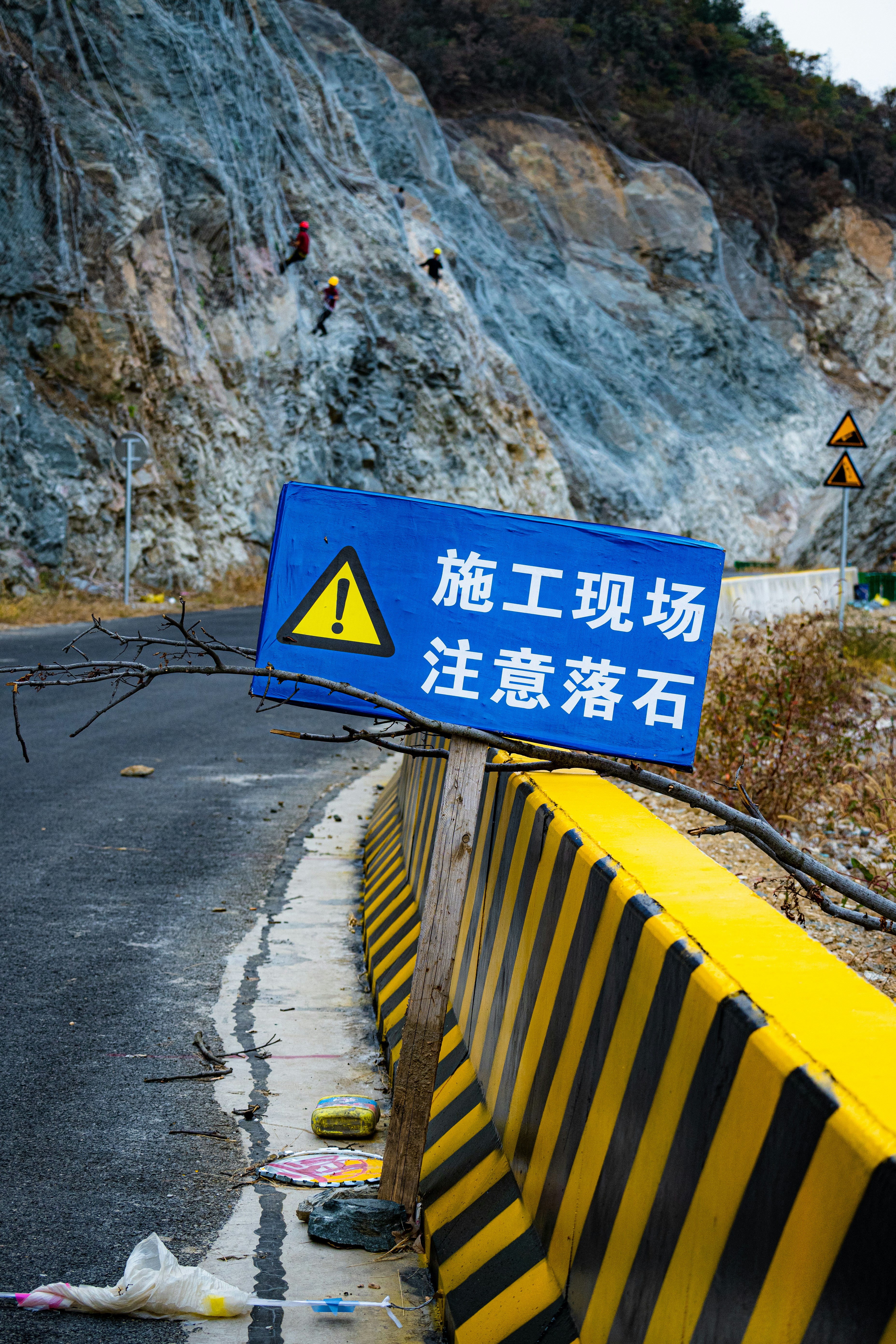 Tilted Blue Warning Sign Over Cliffside Guardrail as Climbers Scale the Weathered Slope