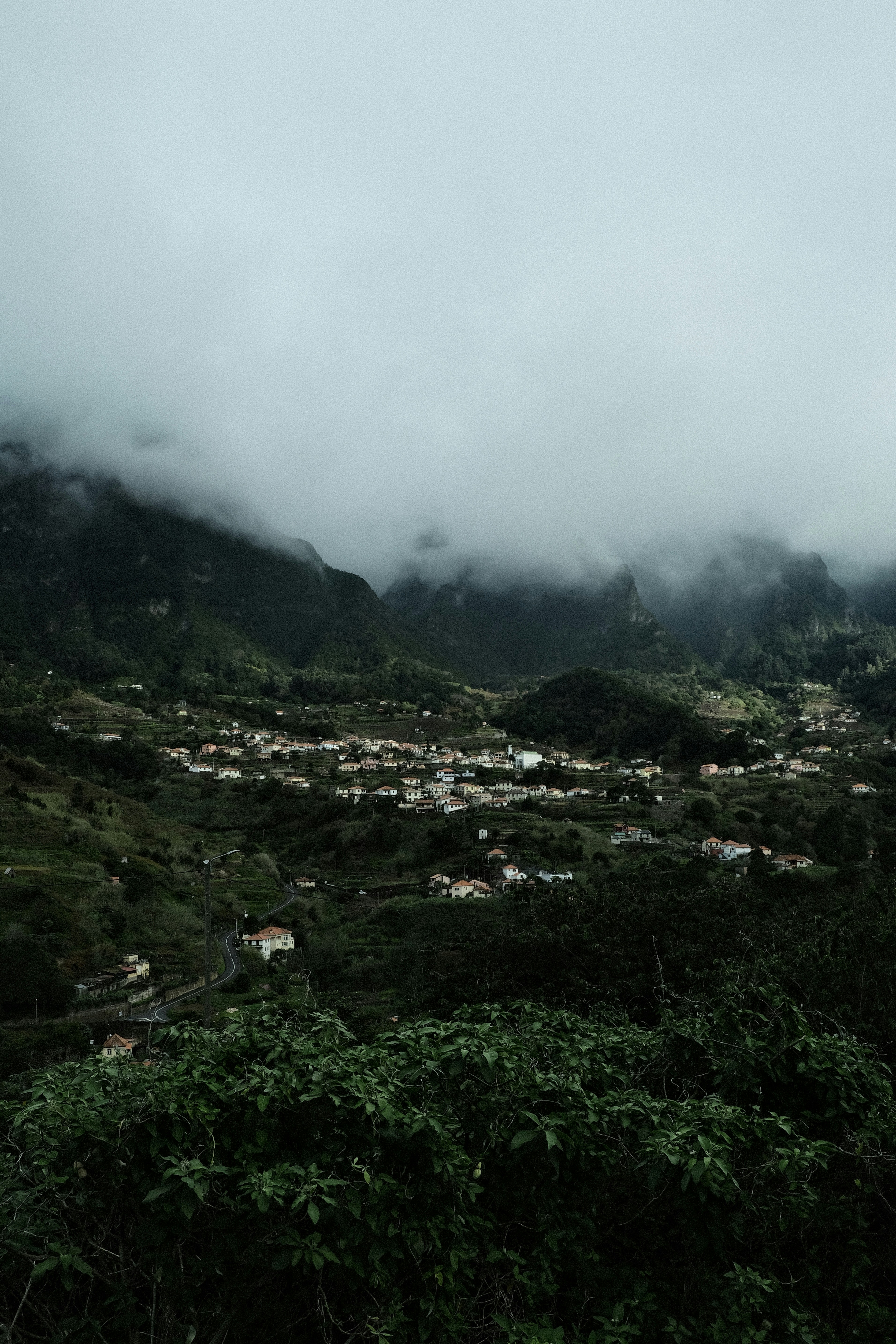 Clouds descend over a lush green valley, partially revealing a small village nestled among rolling hills.