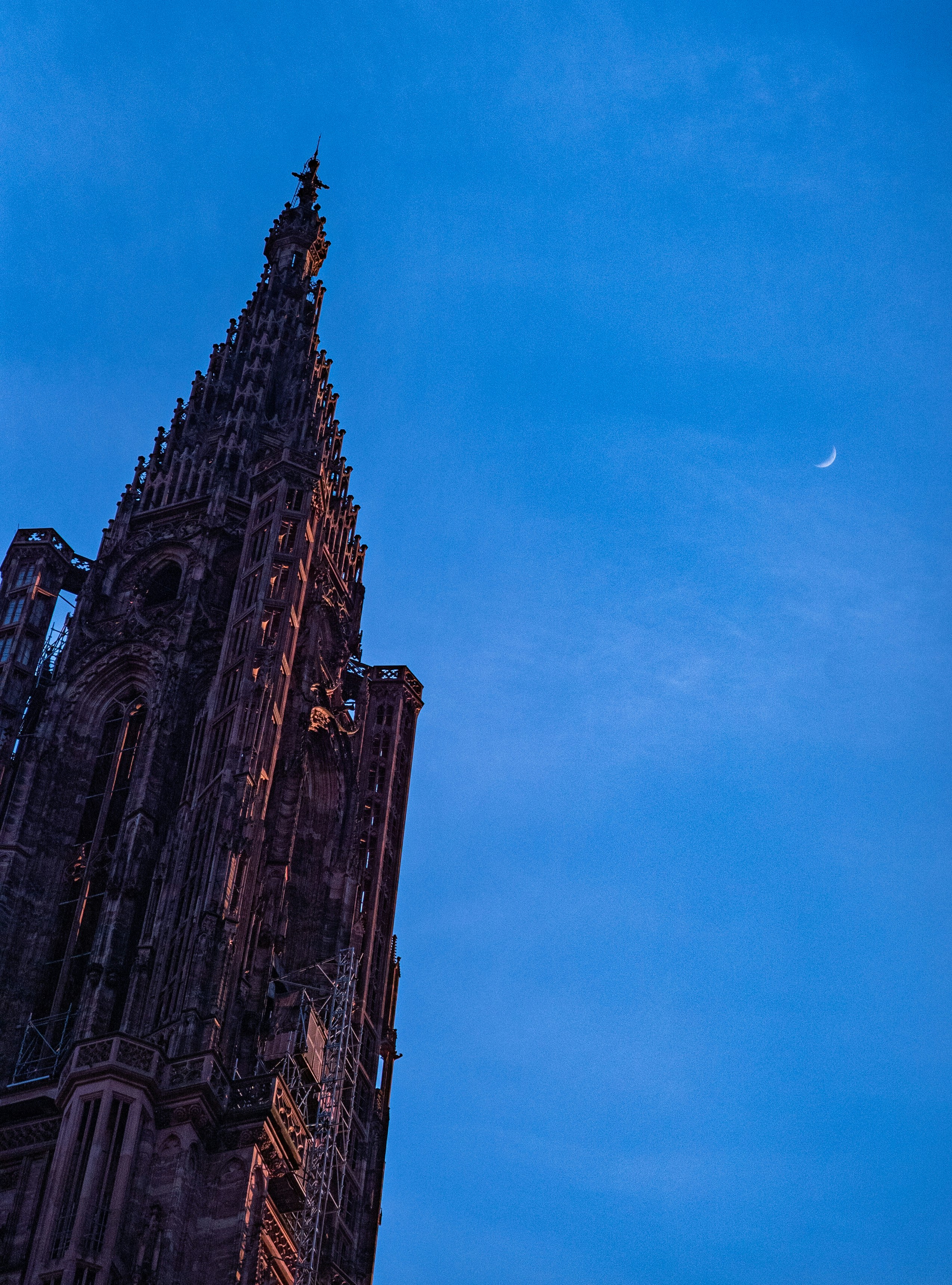Gothic spire of Strasbourg Cathedral against a deep blue evening sky with a crescent moon.