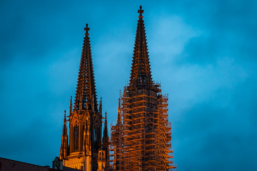 Two Gothic cathedral spires at blue hour, the right spire wrapped in scaffolding and warm illumination against a cyan sky.