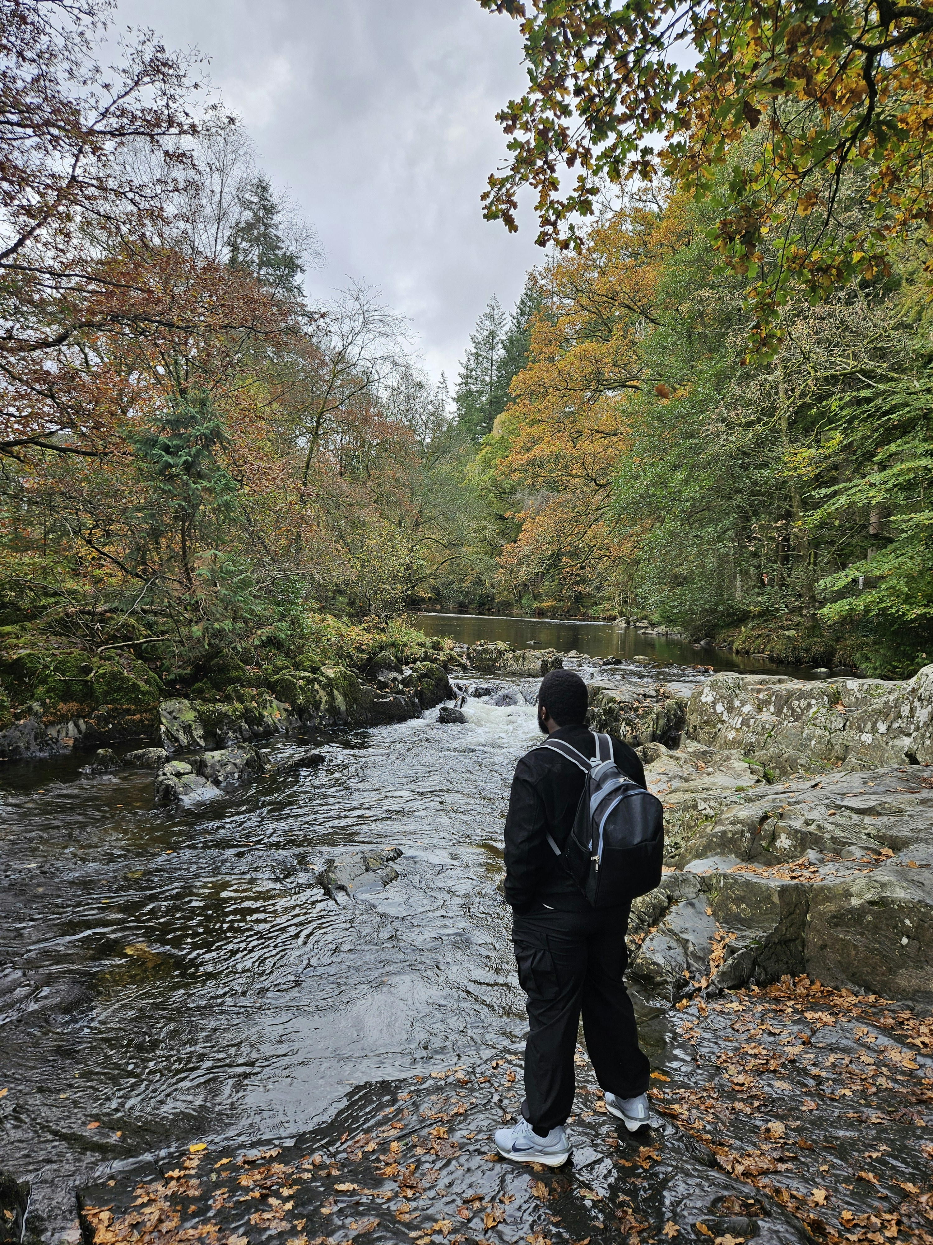 A lone traveler with a backpack stands on wet rocks beside a gently flowing river, framed by autumn trees. This photo captures a moment of quiet exploration in a forested gorge.