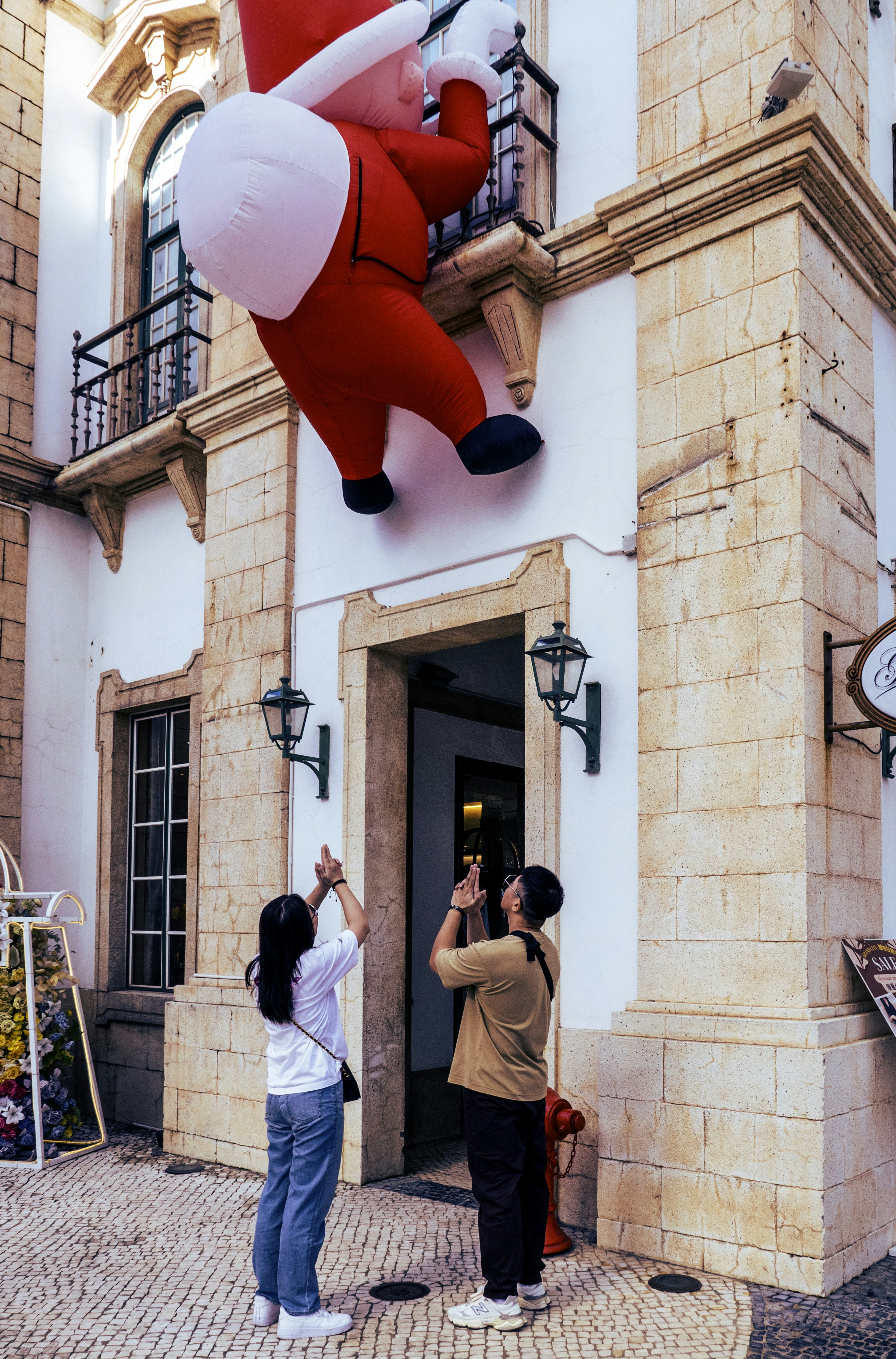 Two visitors raise their phones to photograph a colossal inflatable Santa Claus dangling from a balcony above a stone arcade, a festive scene unfolding beneath.