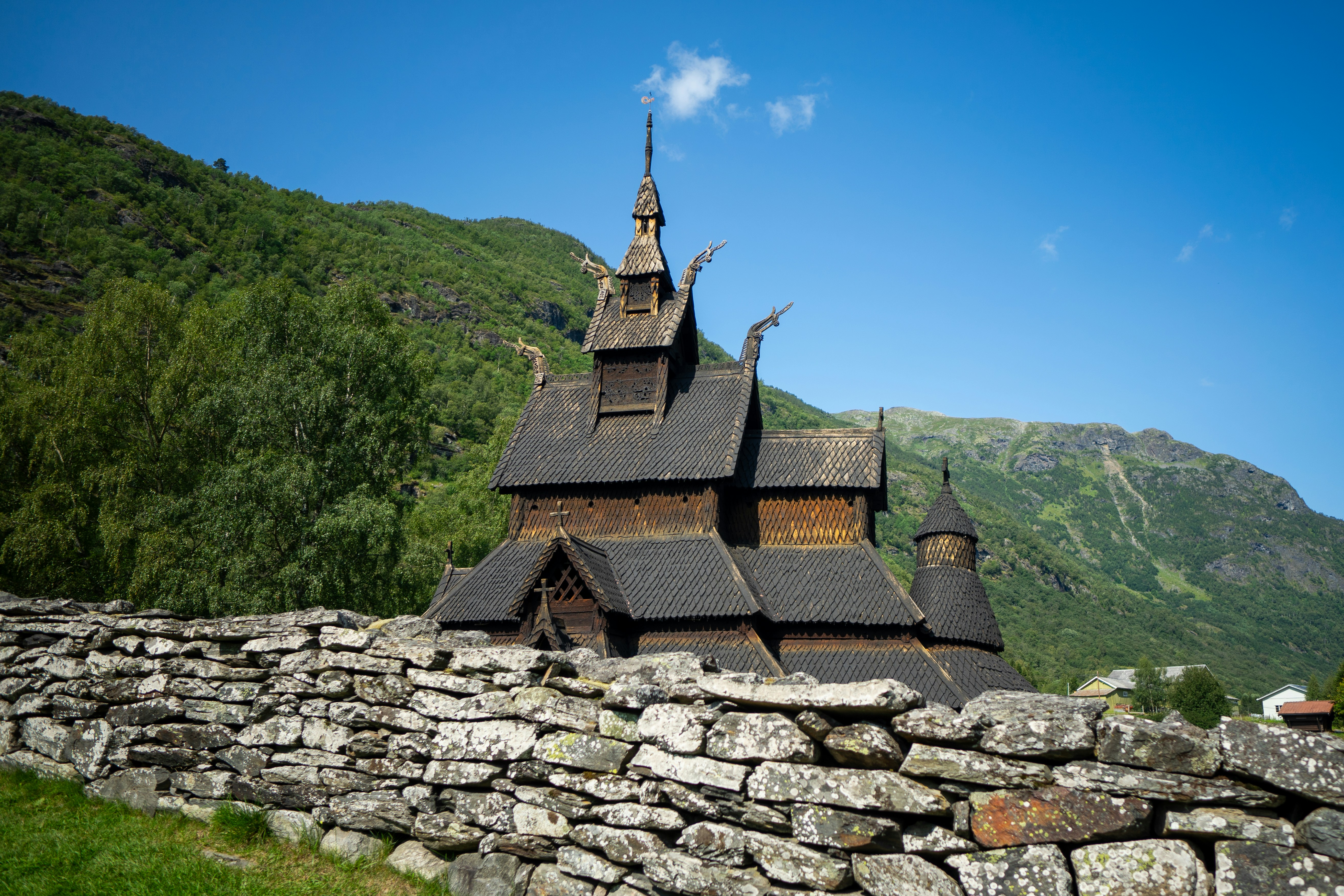 A traditional stave church stands proudly against a backdrop of lush mountains, framed by a rustic stone wall. The intricate architecture showcases historical craftsmanship.