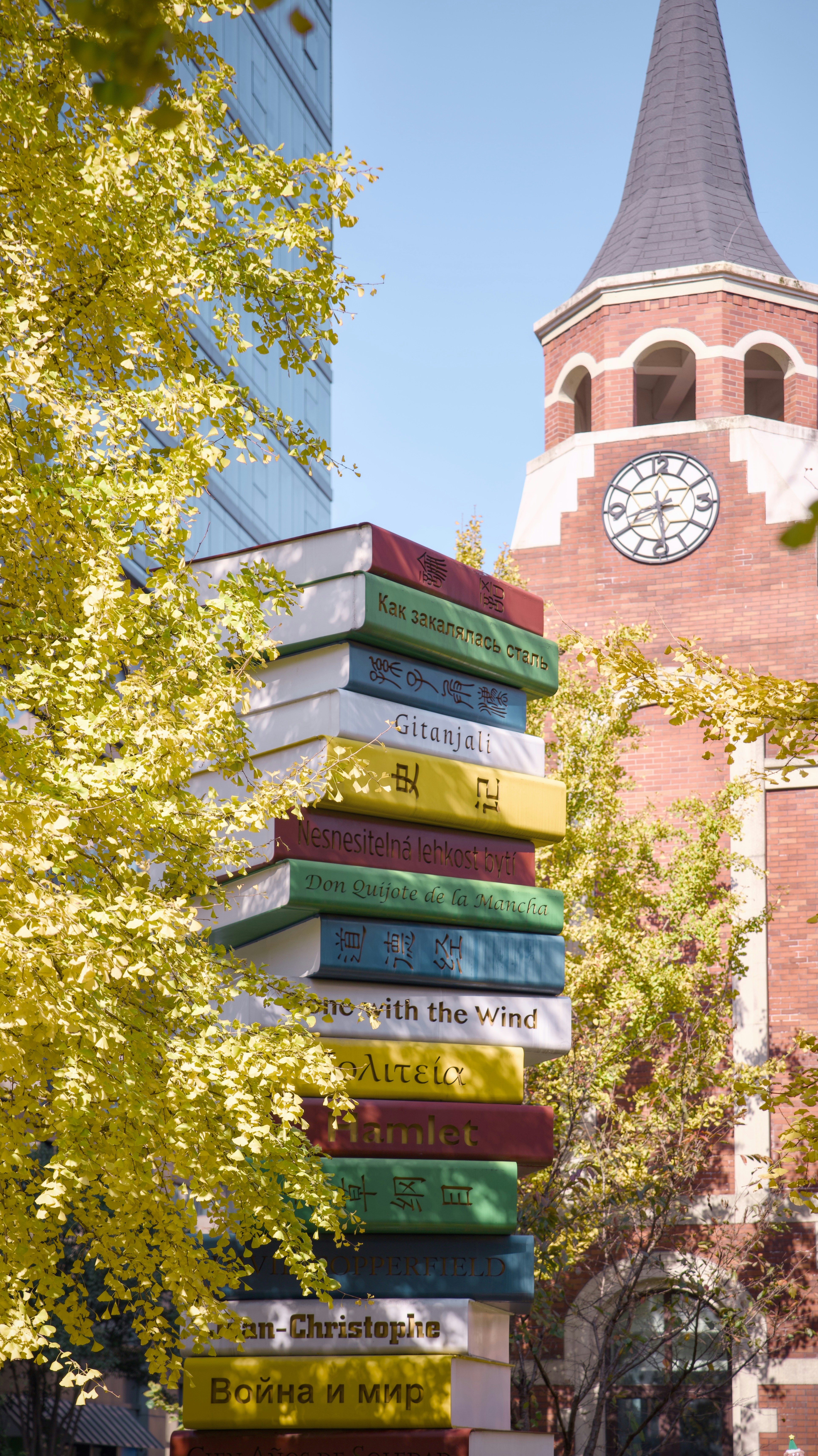 A striking sculpture of stacked books featuring world literature classics in multiple languages stands against the backdrop of a brick church tower with a clock. The composition is enhanced by golden autumn foliage, creating a perfect blend of education, architecture, and nature.