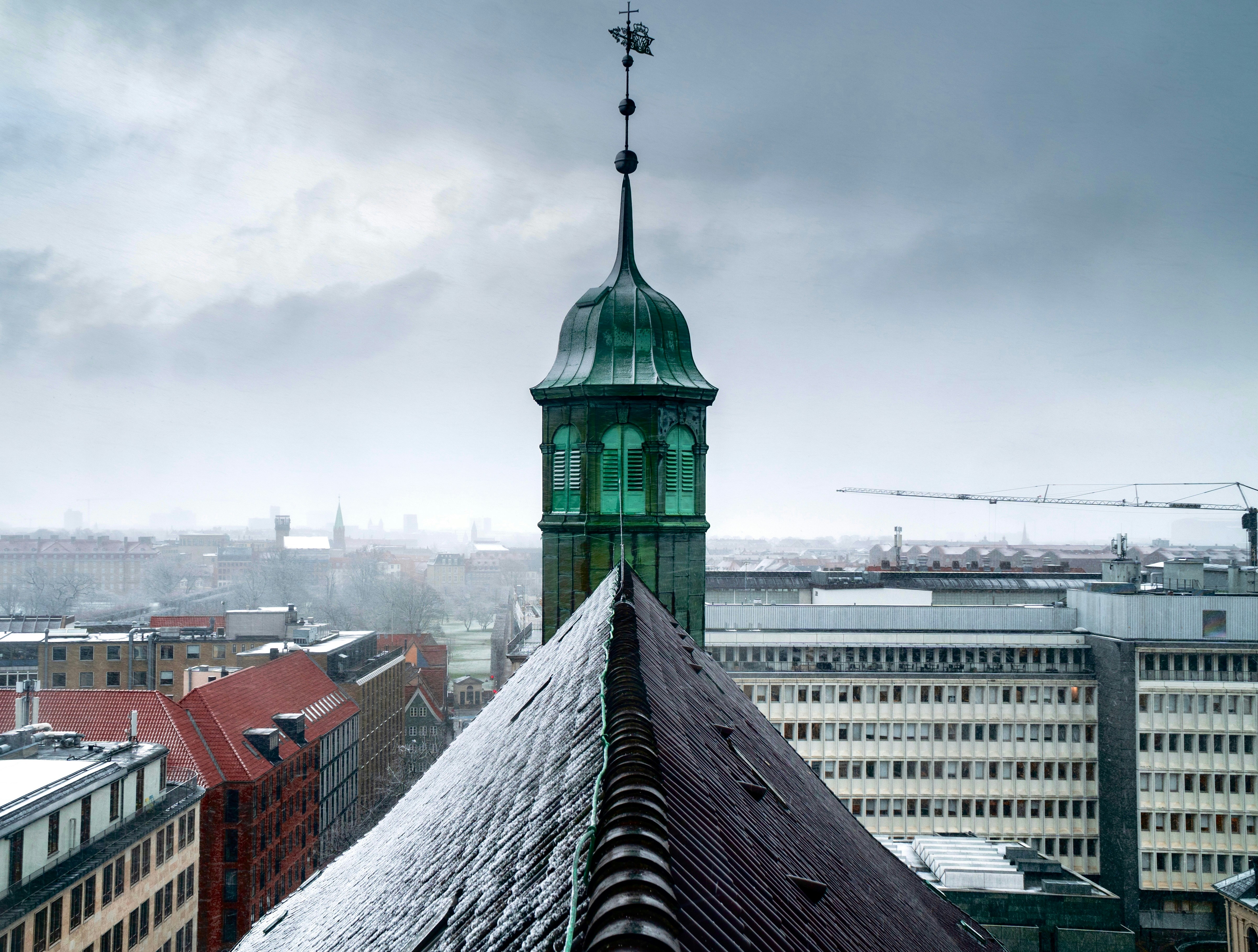 Snow-dusted rooftop and green spire overlook a cityscape under a cloudy sky.
