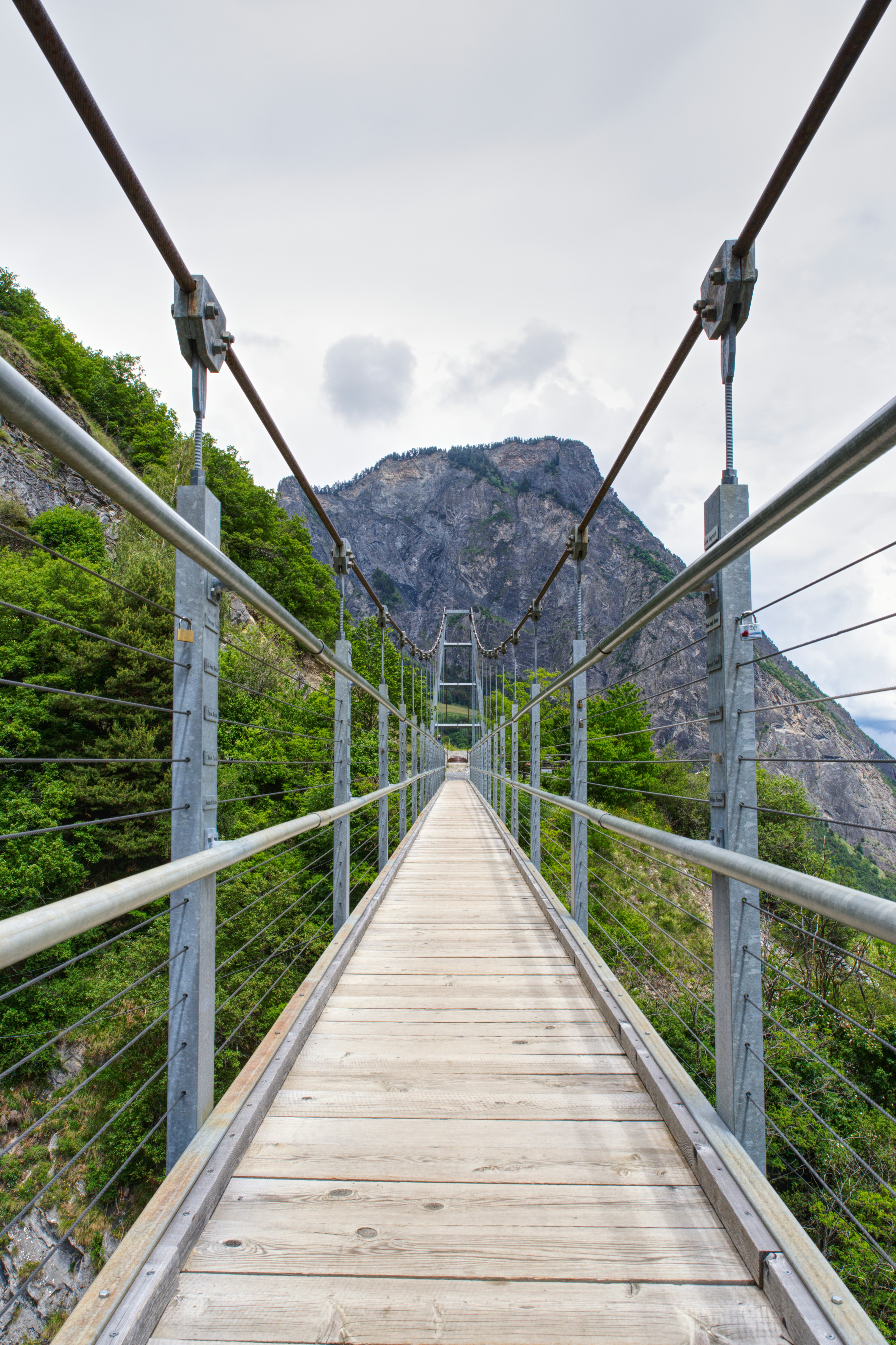 Suspension bridge stretching across a lush valley, flanked by towering mountains and vibrant greenery. The pathway invites exploration.