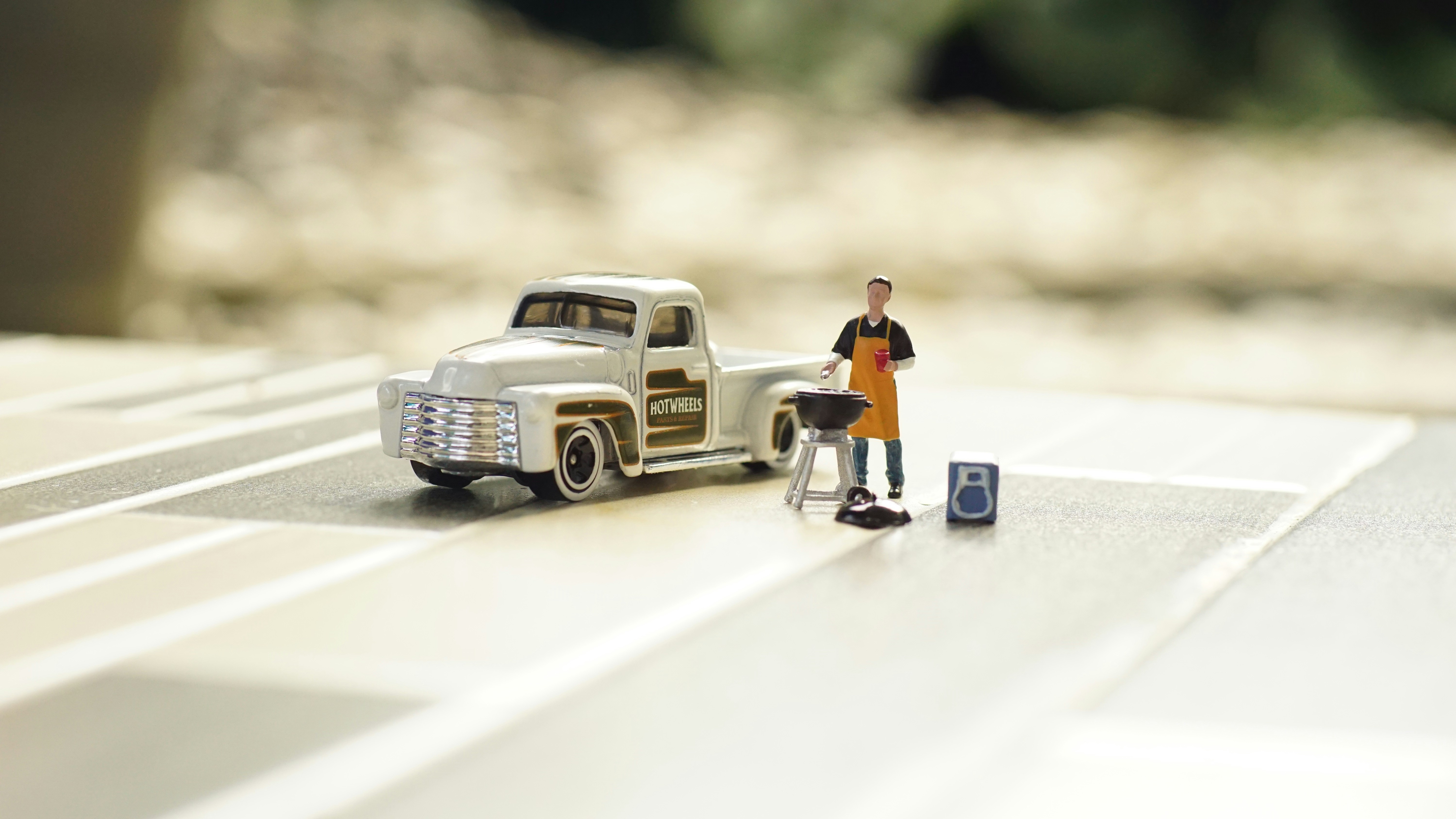 A tiny vintage pickup truck parked beside a miniature barbecue setup, with a figure in an apron preparing food. The scene captures a playful outdoor grilling moment.