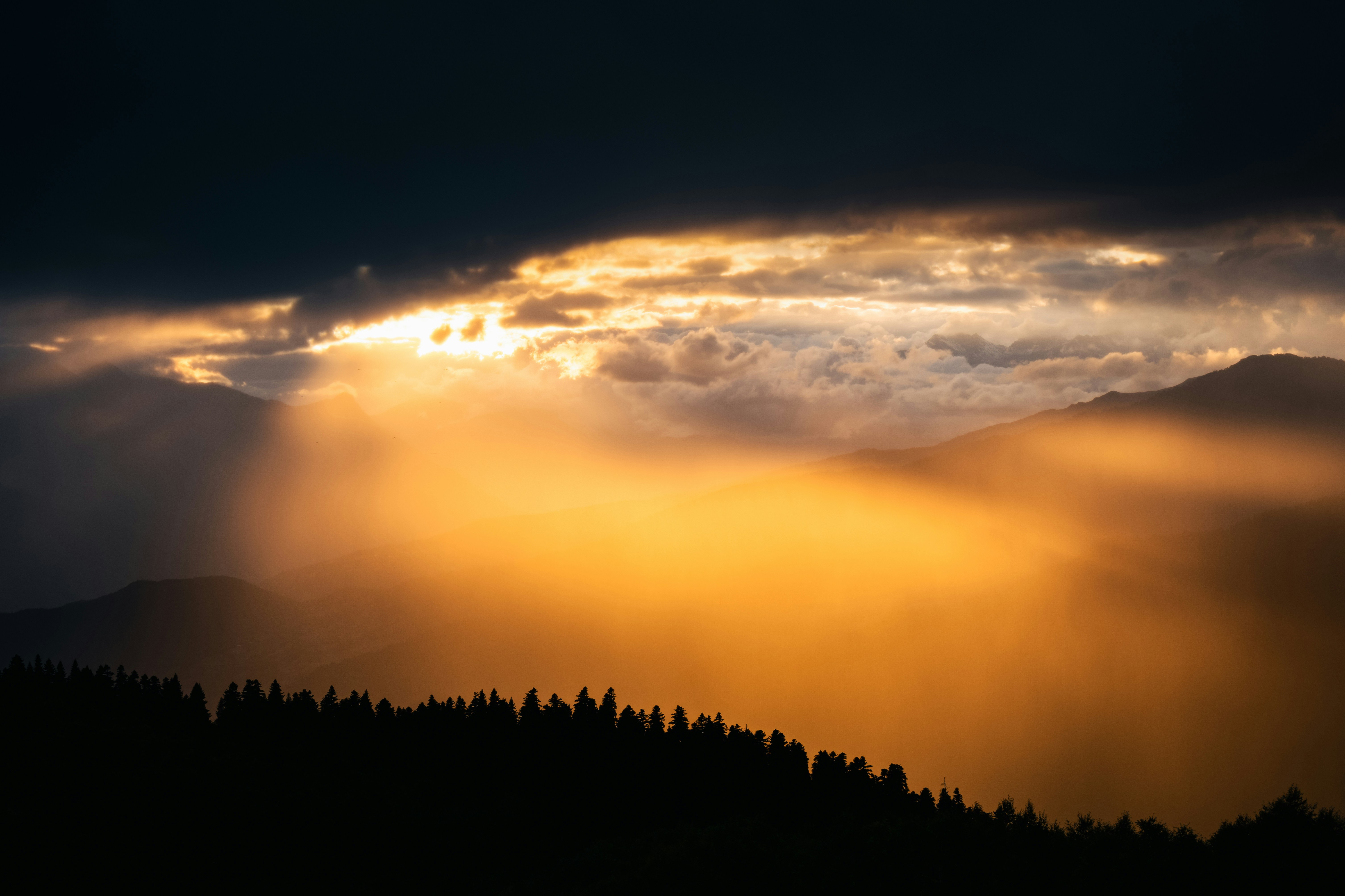 Sun rays pierce through dramatic clouds over silhouetted mountains.
